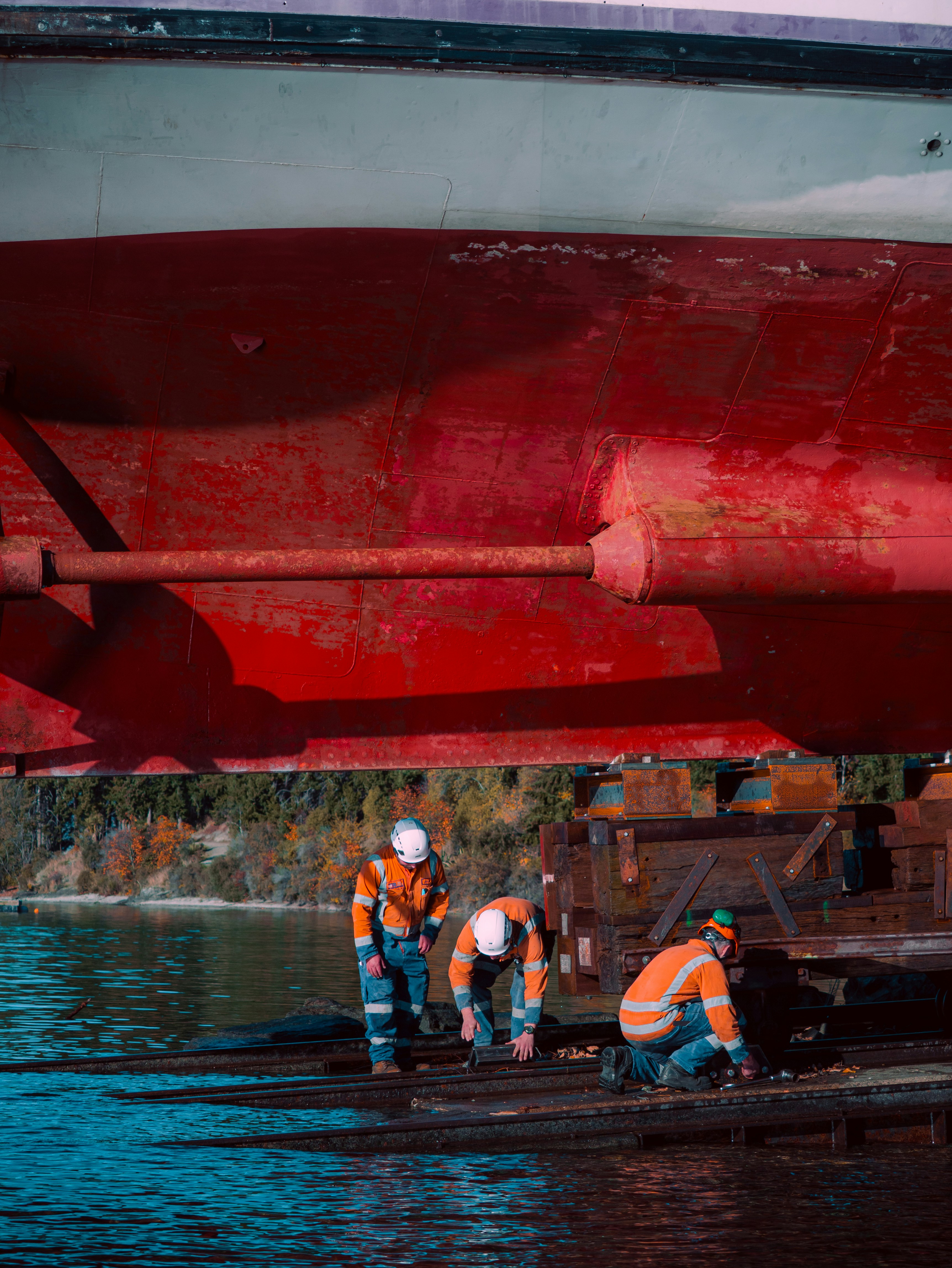 Crew at the keel: workers under a red vessel in a river dockyard