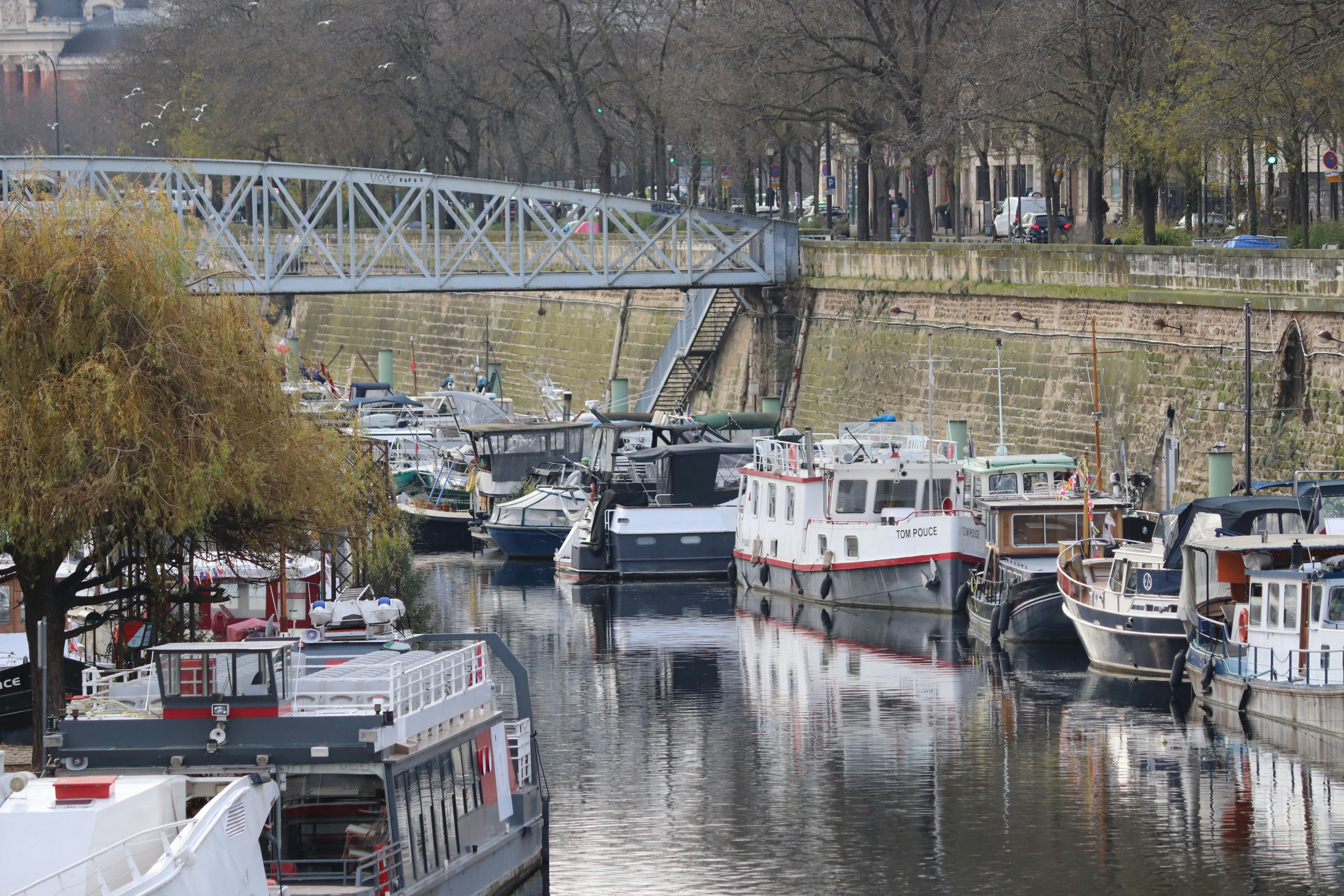 A tranquil canal scene featuring various boats lined along the waterway, with a bridge in the background and trees lining the banks.