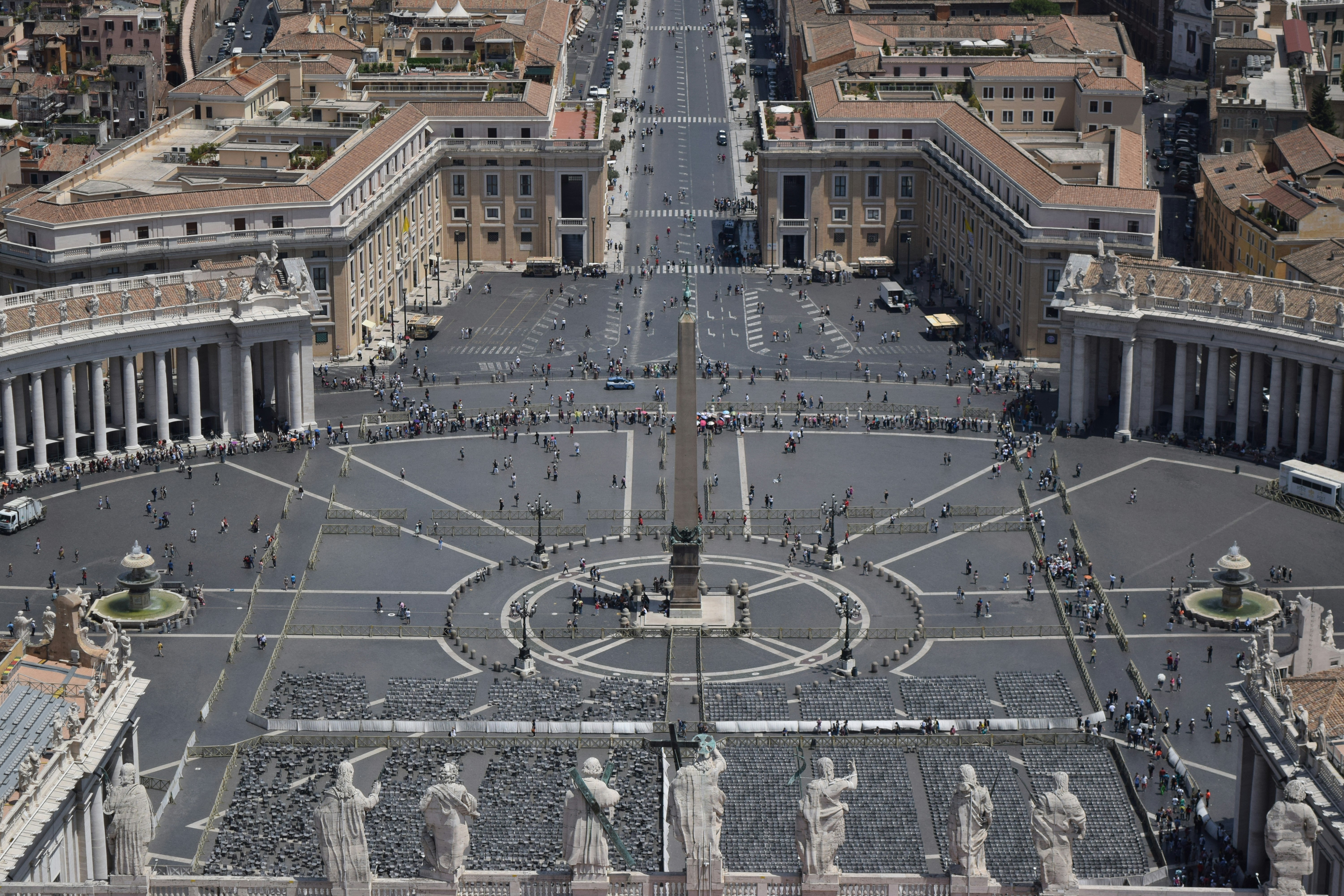 Bird's-eye view of St. Peter's Square, showcasing the iconic architecture and bustling activity of visitors. The central obelisk stands as a focal point amidst the expansive plaza.