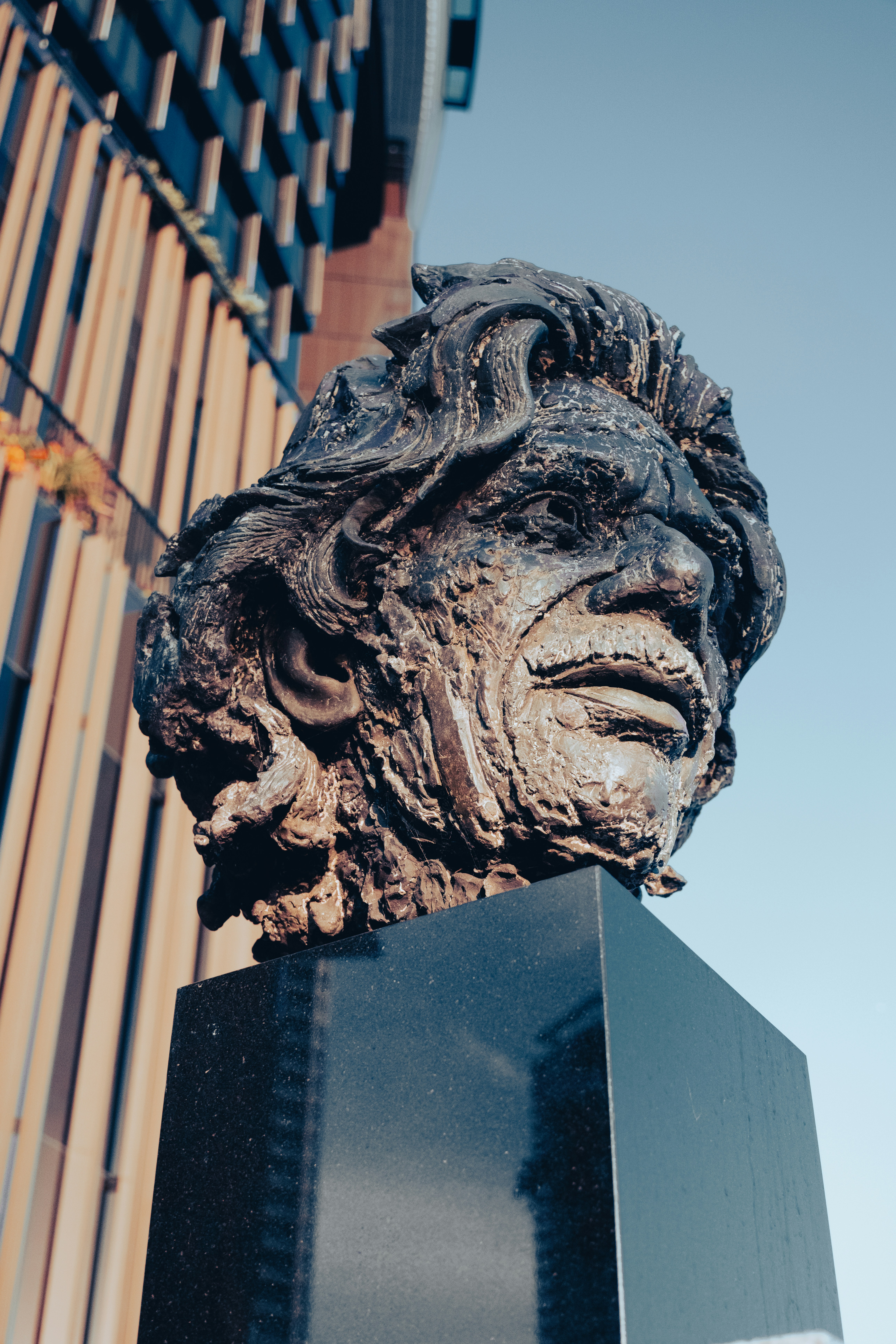 Detailed bronze sculpture of a man's head, showcasing expressive features and textured hair, set against a modern architectural backdrop.