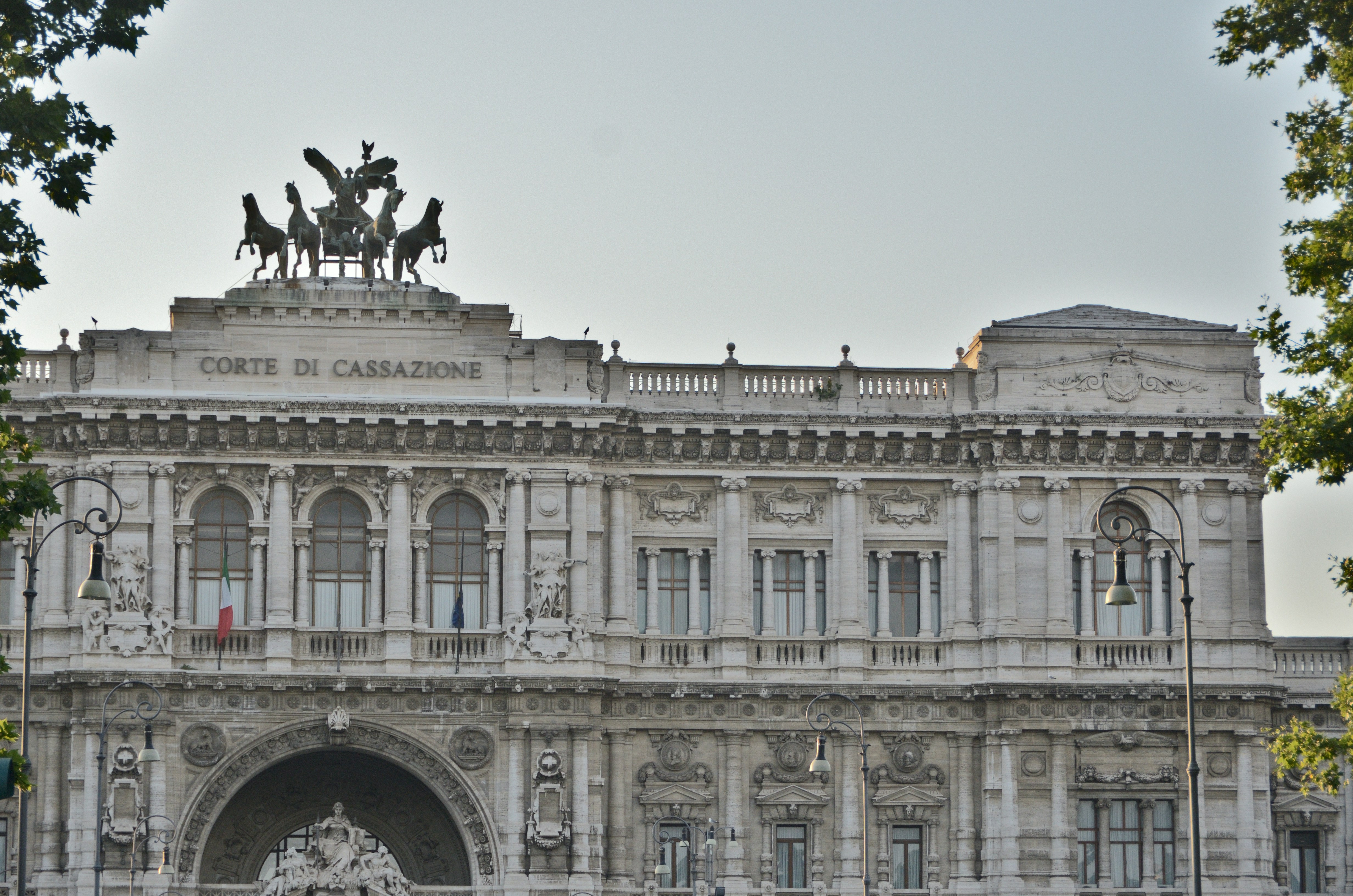 The grand facade of Corte di Cassazione, featuring intricate architectural details and a prominent sculpture atop. Flags flutter gently in the breeze.