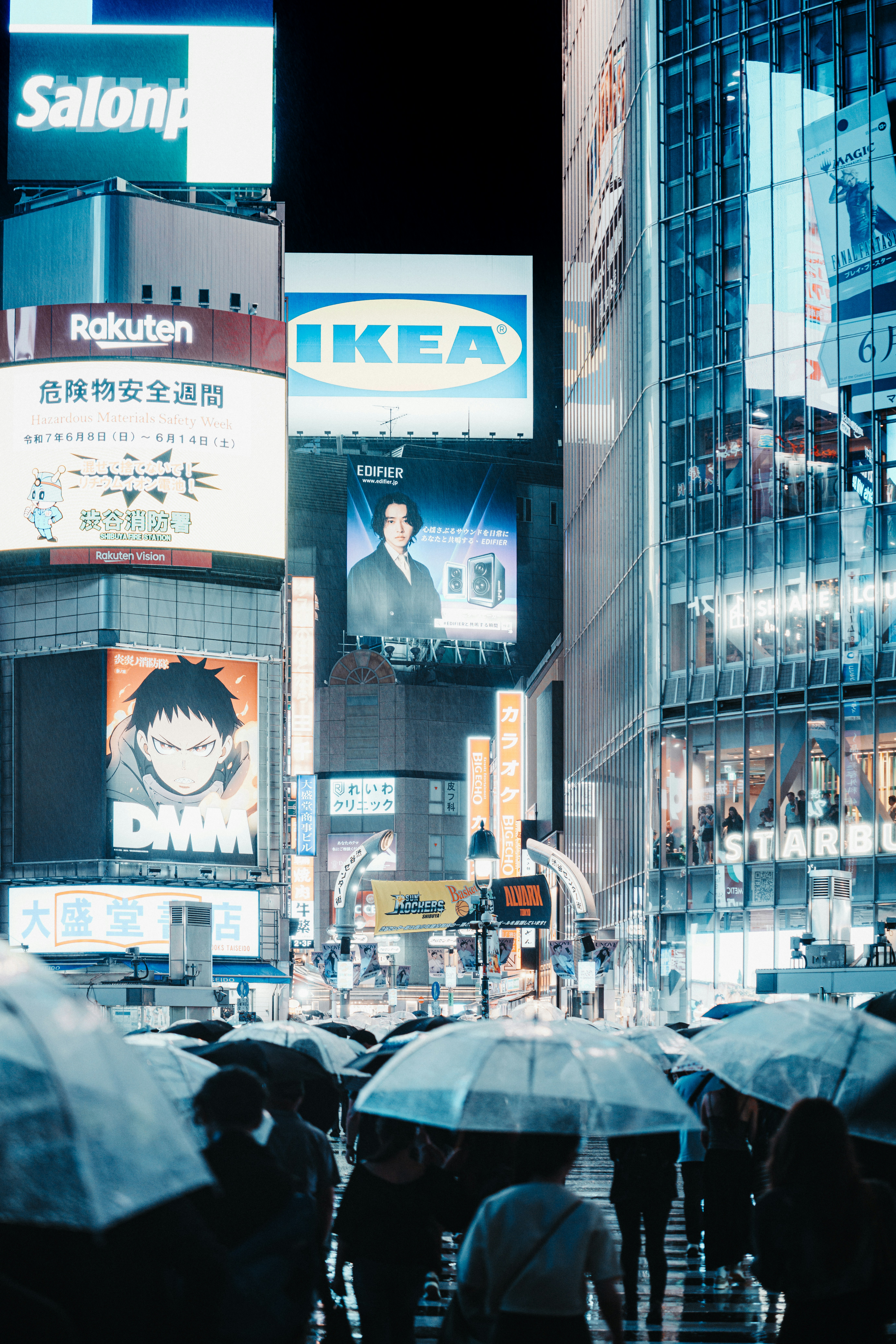 Crowd of people with umbrellas crossing a bustling city intersection illuminated by vibrant neon advertisements and digital billboards.