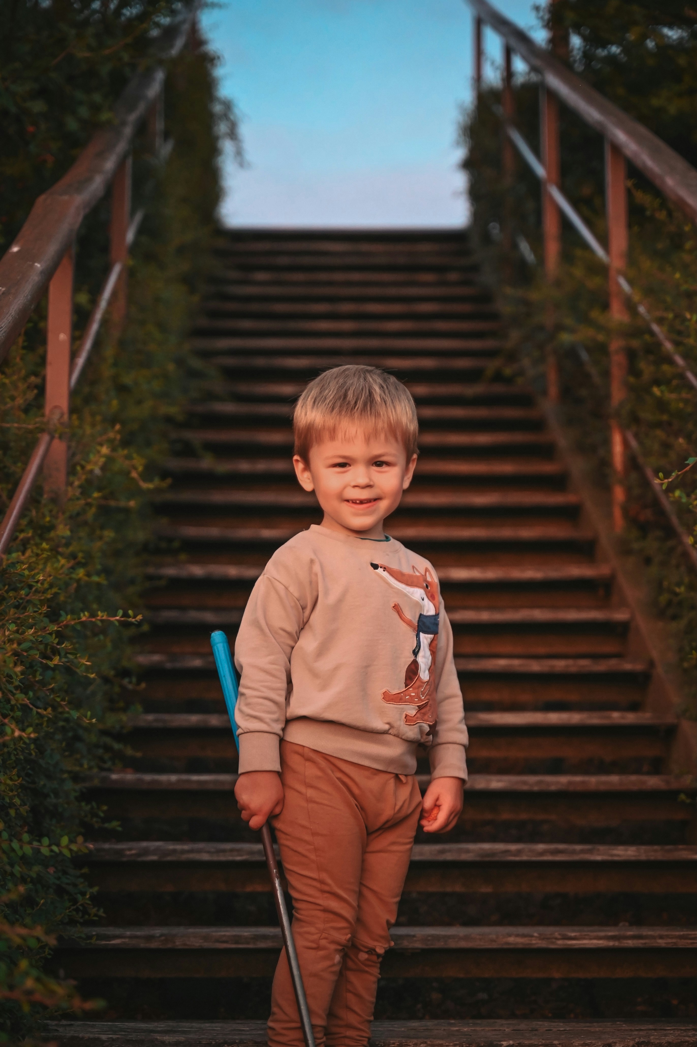 Young child smiling on a staircase surrounded by greenery, holding a stick. The scene evokes a sense of playfulness and exploration.