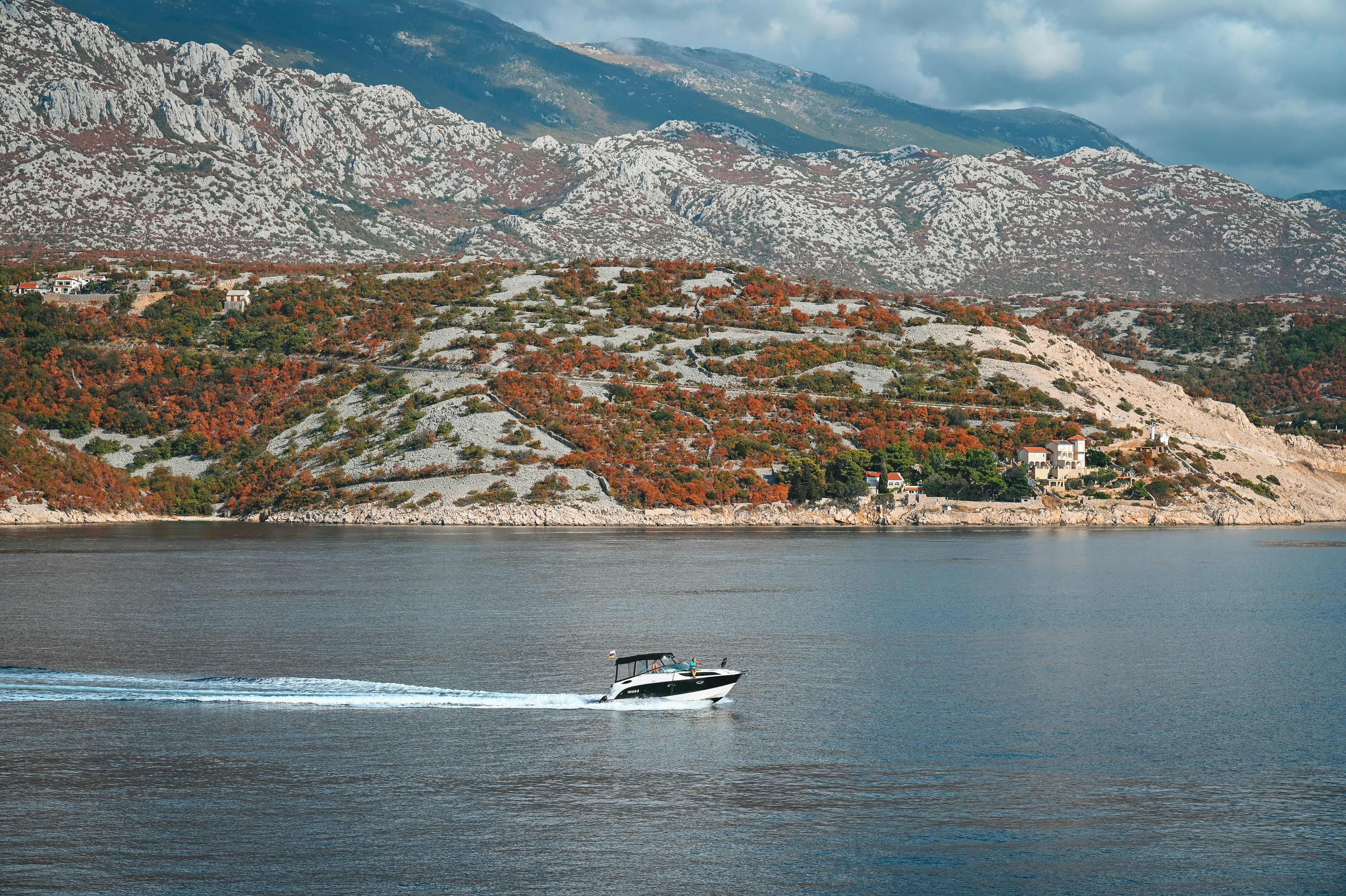A sleek boat glides through calm waters, framed by rugged coastal hills adorned with autumn foliage.