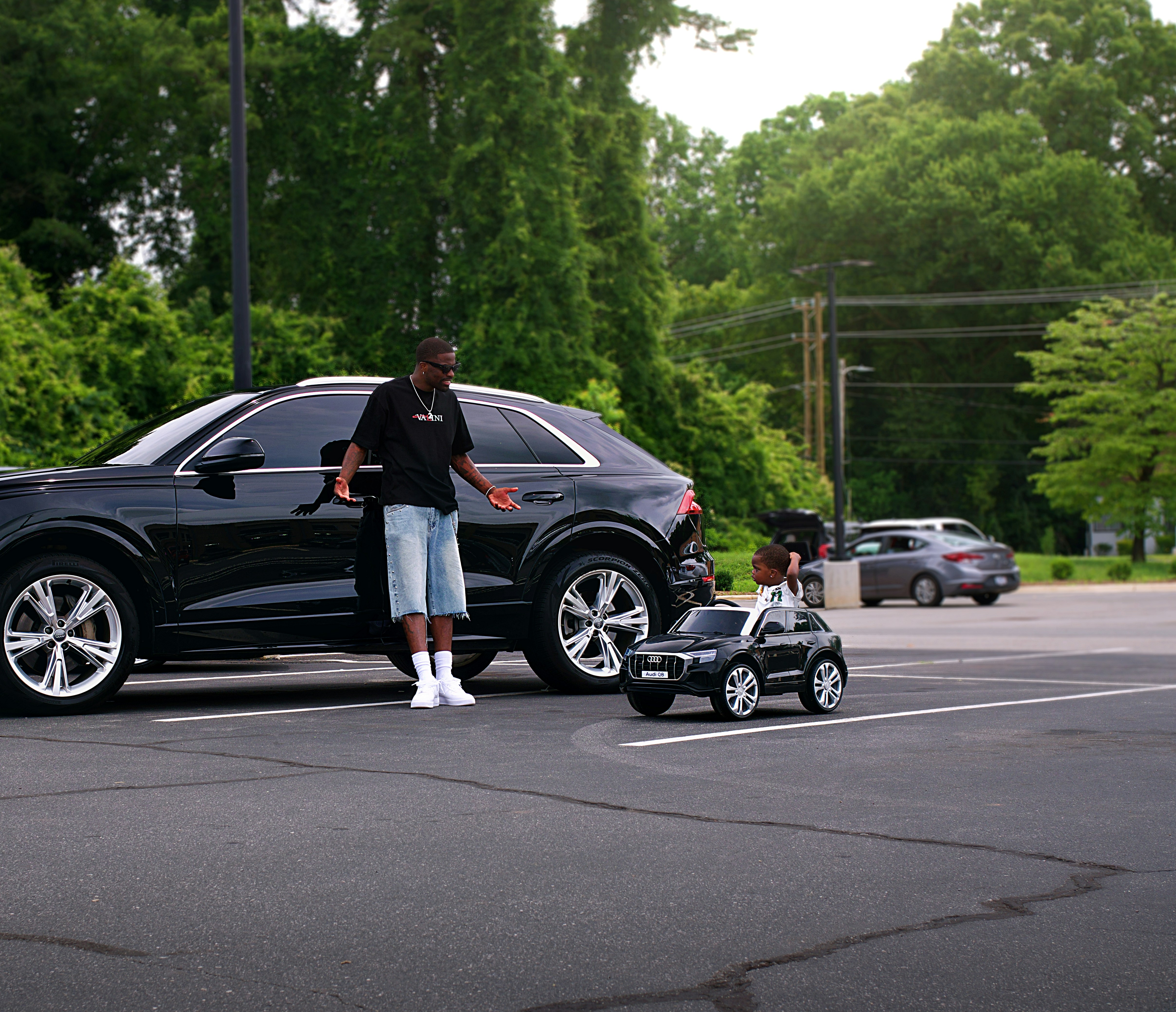 A man interacts with a child in a toy car beside a sleek black SUV in a parking lot, surrounded by lush greenery.