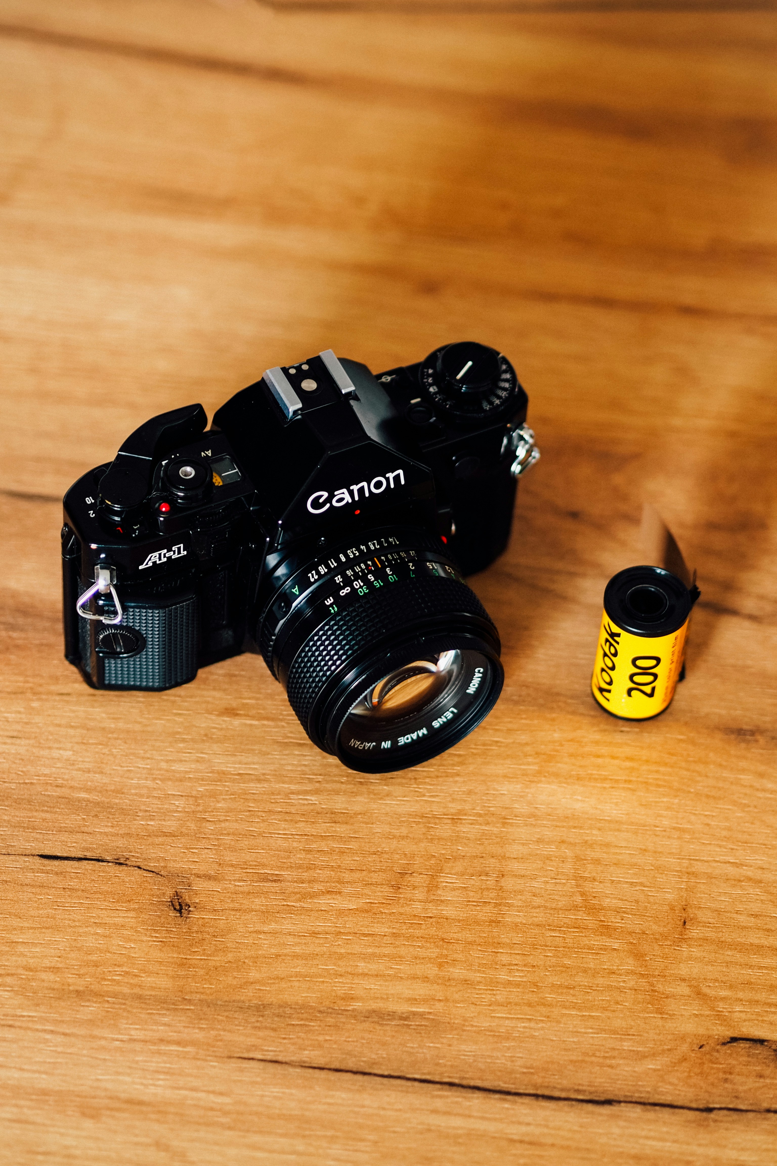 Canon film camera positioned on a wooden surface with a roll of Kodak 200 film beside it.