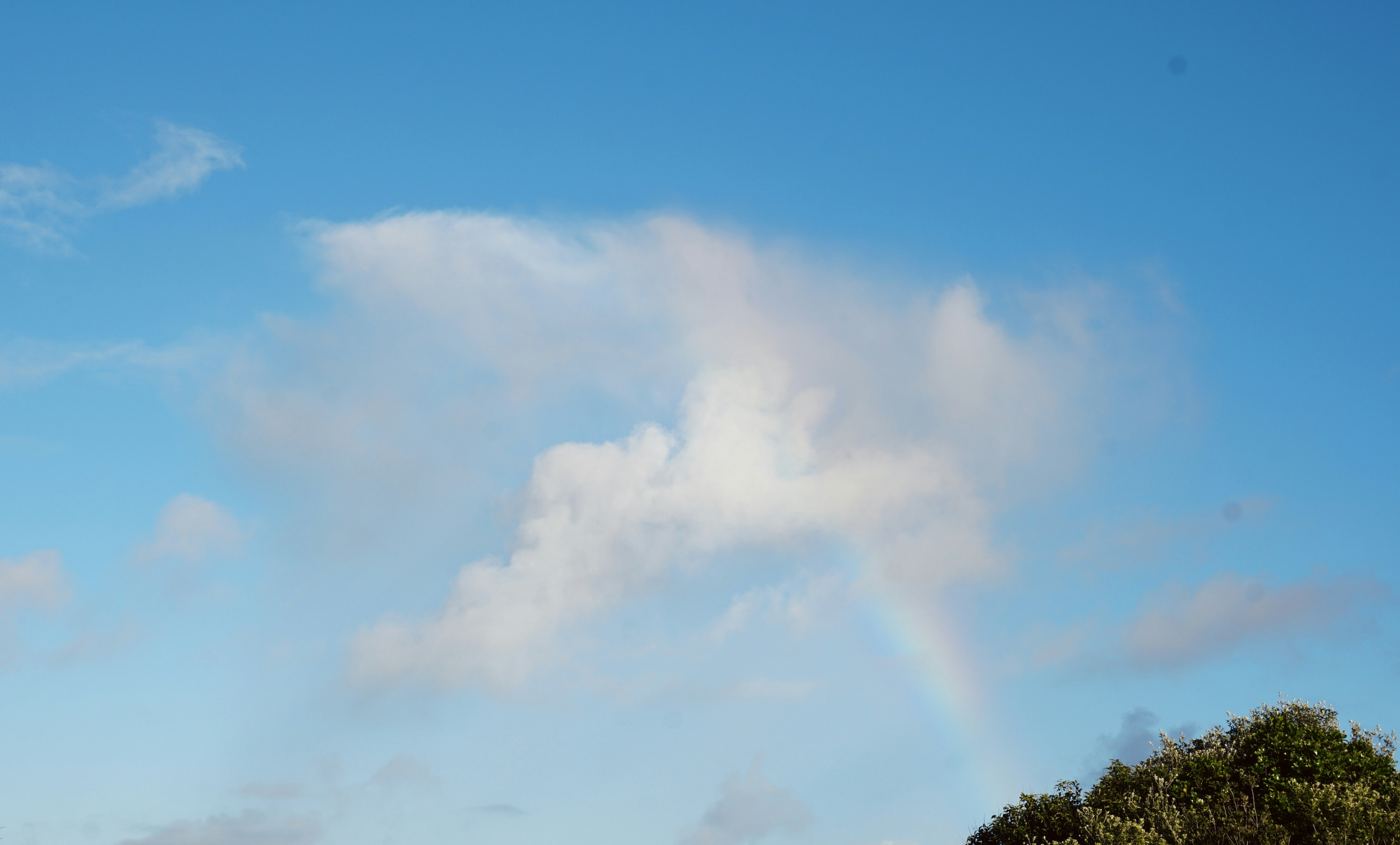 A delicate rainbow arcs through a backdrop of soft clouds against a clear blue sky.