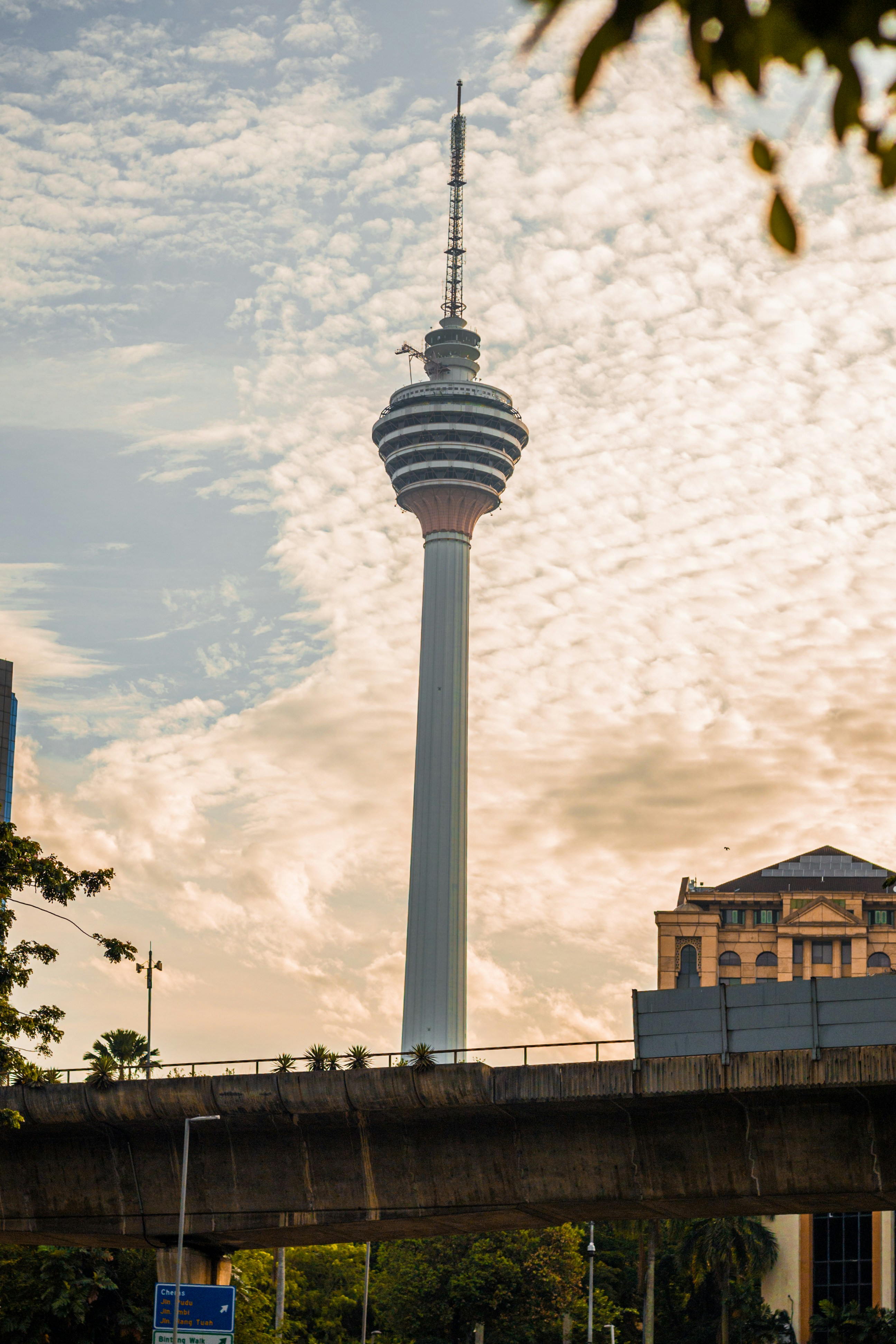 Telecommunications tower rising against a backdrop of wispy clouds during sunset, framed by urban architecture and greenery.