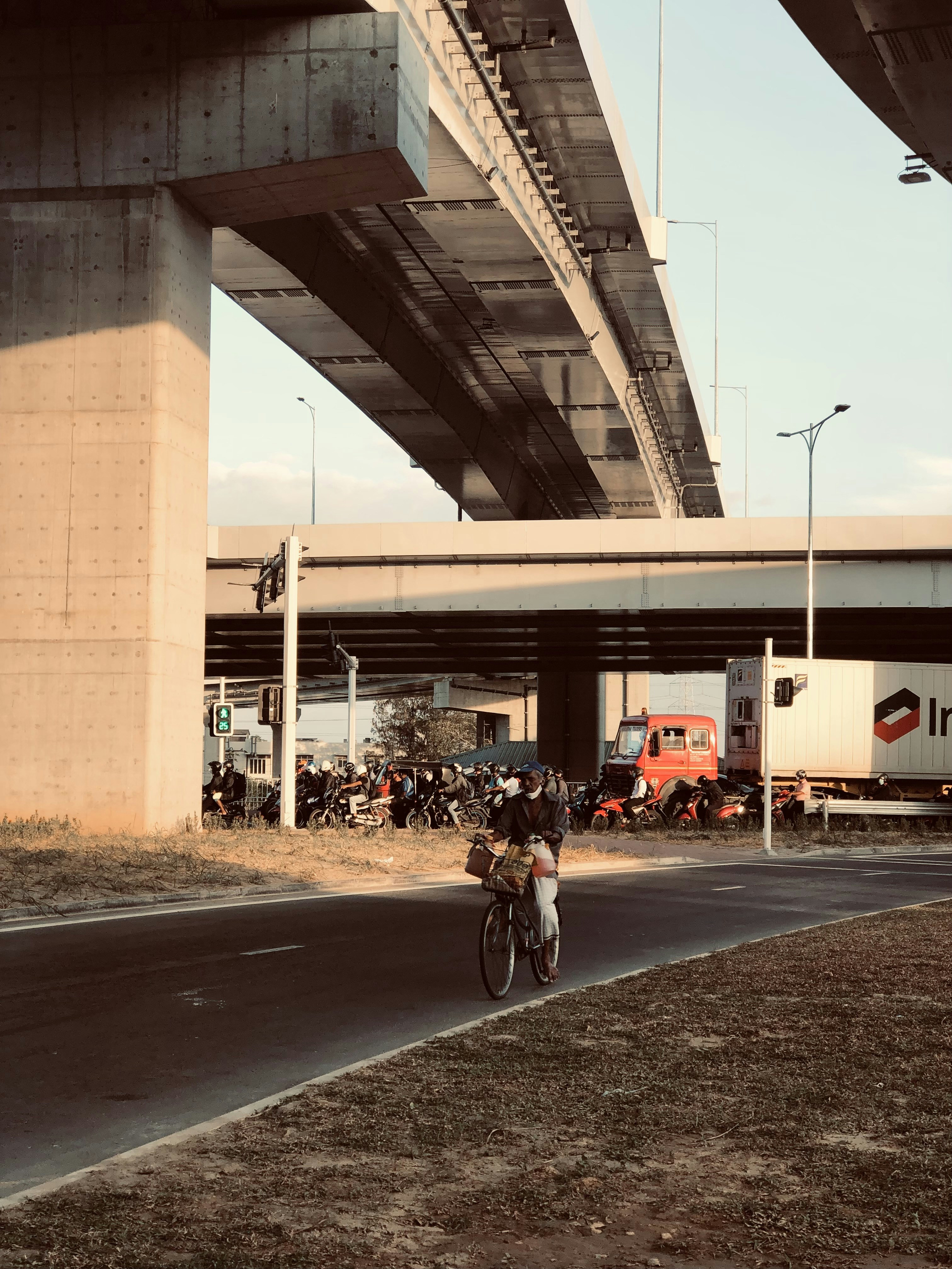Amidst the chaos of a heavily trafficked Sri Lankan road, a cyclist threads his way forward. | A cyclist rides underneath an overpass.