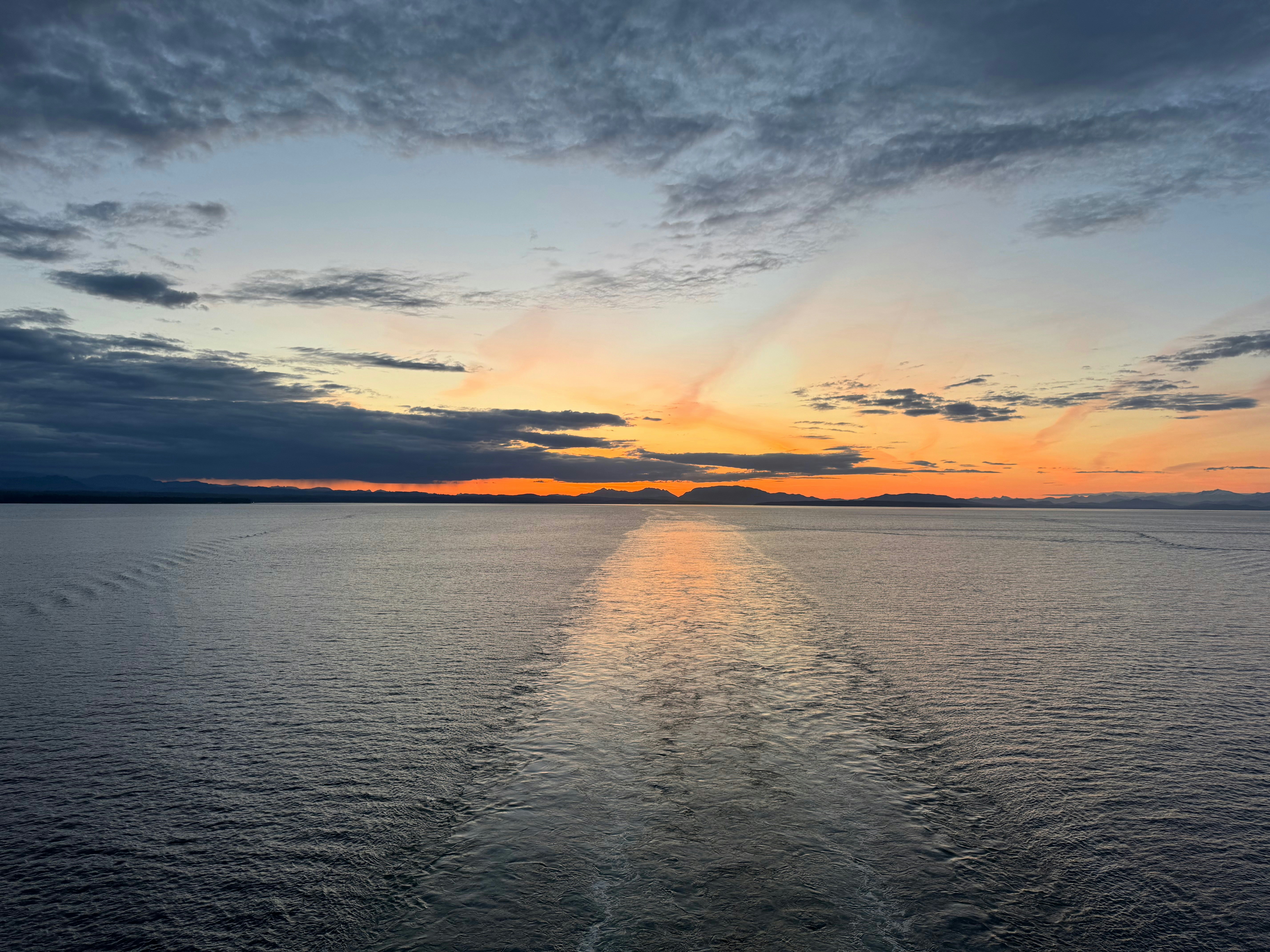 Vibrant sunset reflecting on calm waters, with distant mountains silhouetted against the colorful sky.