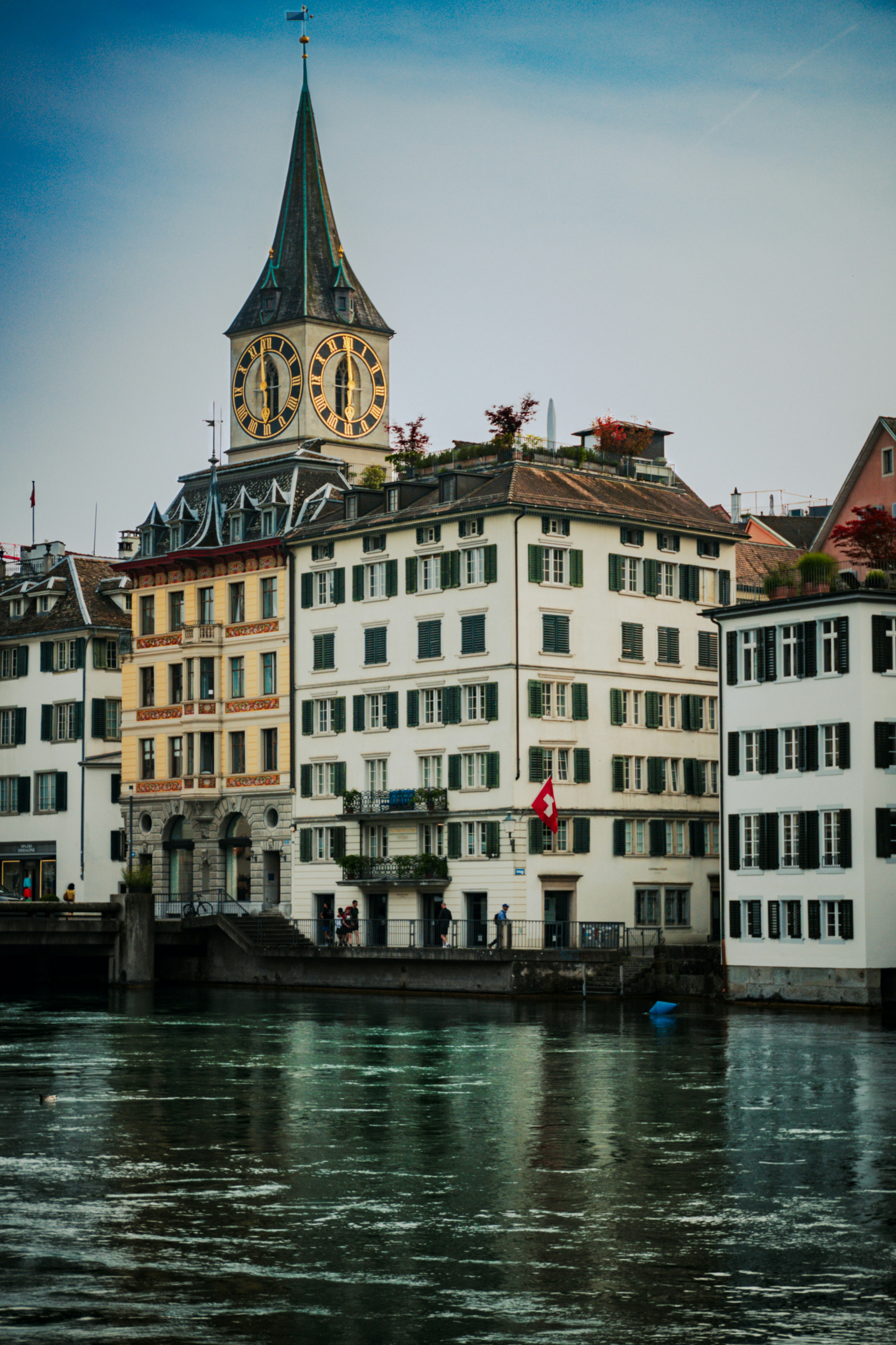 Historic clock tower stands tall amidst charming buildings along the riverbank, reflecting the essence of Swiss architecture.