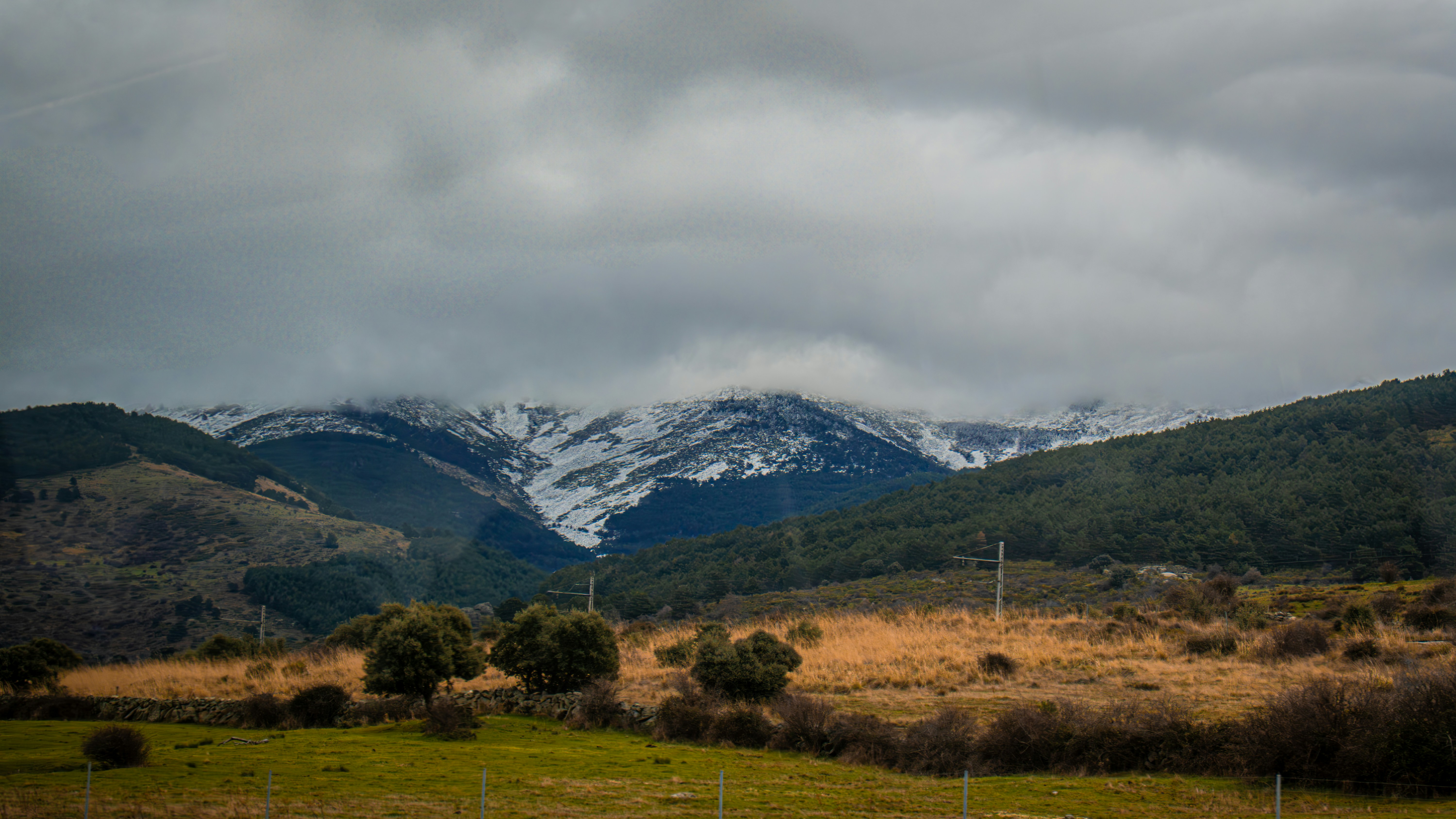 Snow-capped mountains loom under a blanket of clouds, framed by rolling hills and sparse vegetation. A serene landscape captures the essence of nature's quiet beauty.