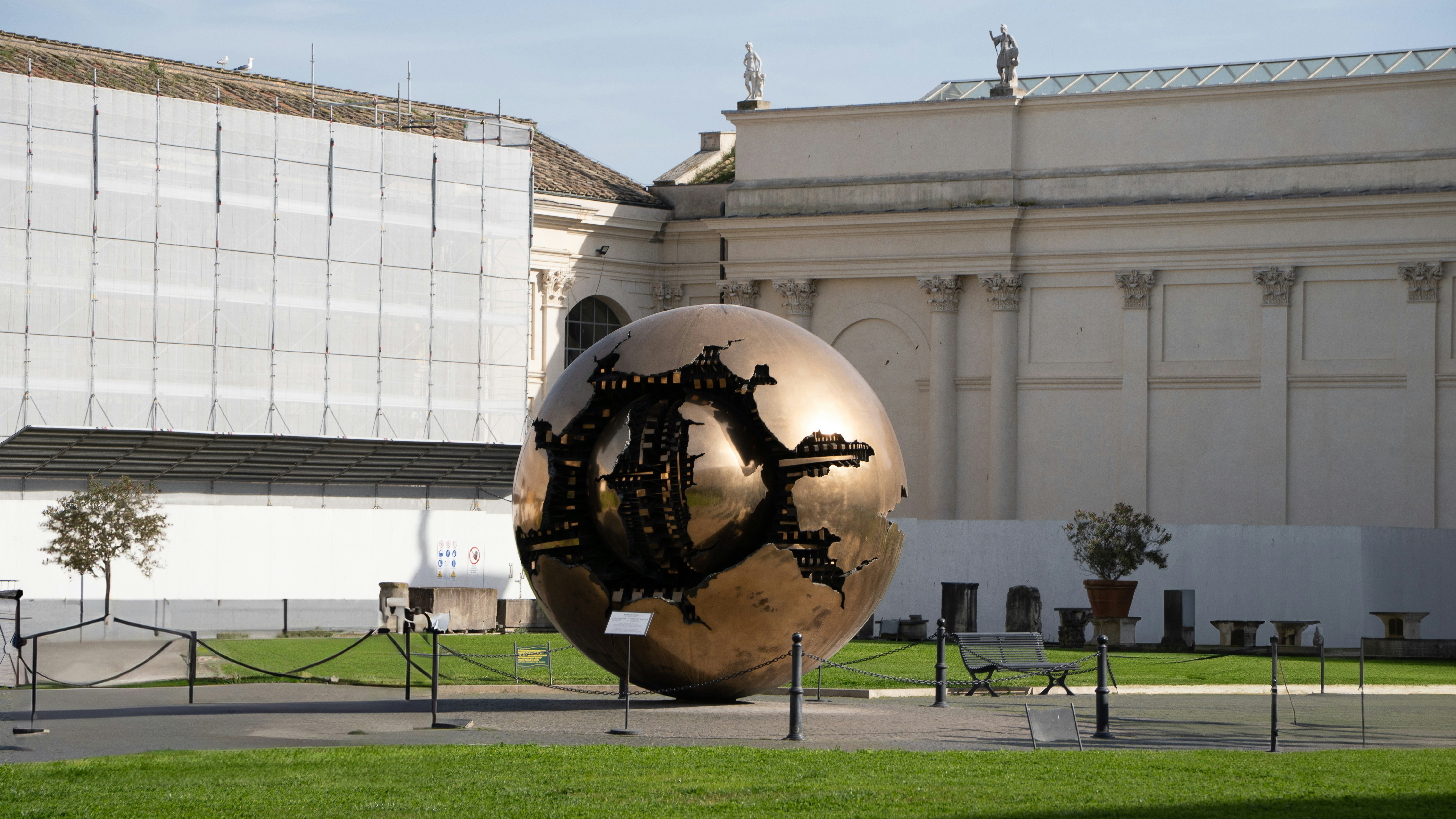 A large metallic sphere featuring intricate designs stands prominently in a landscaped area, surrounded by a historic building and scaffolding. The artwork invites contemplation on the interplay between art and architecture.