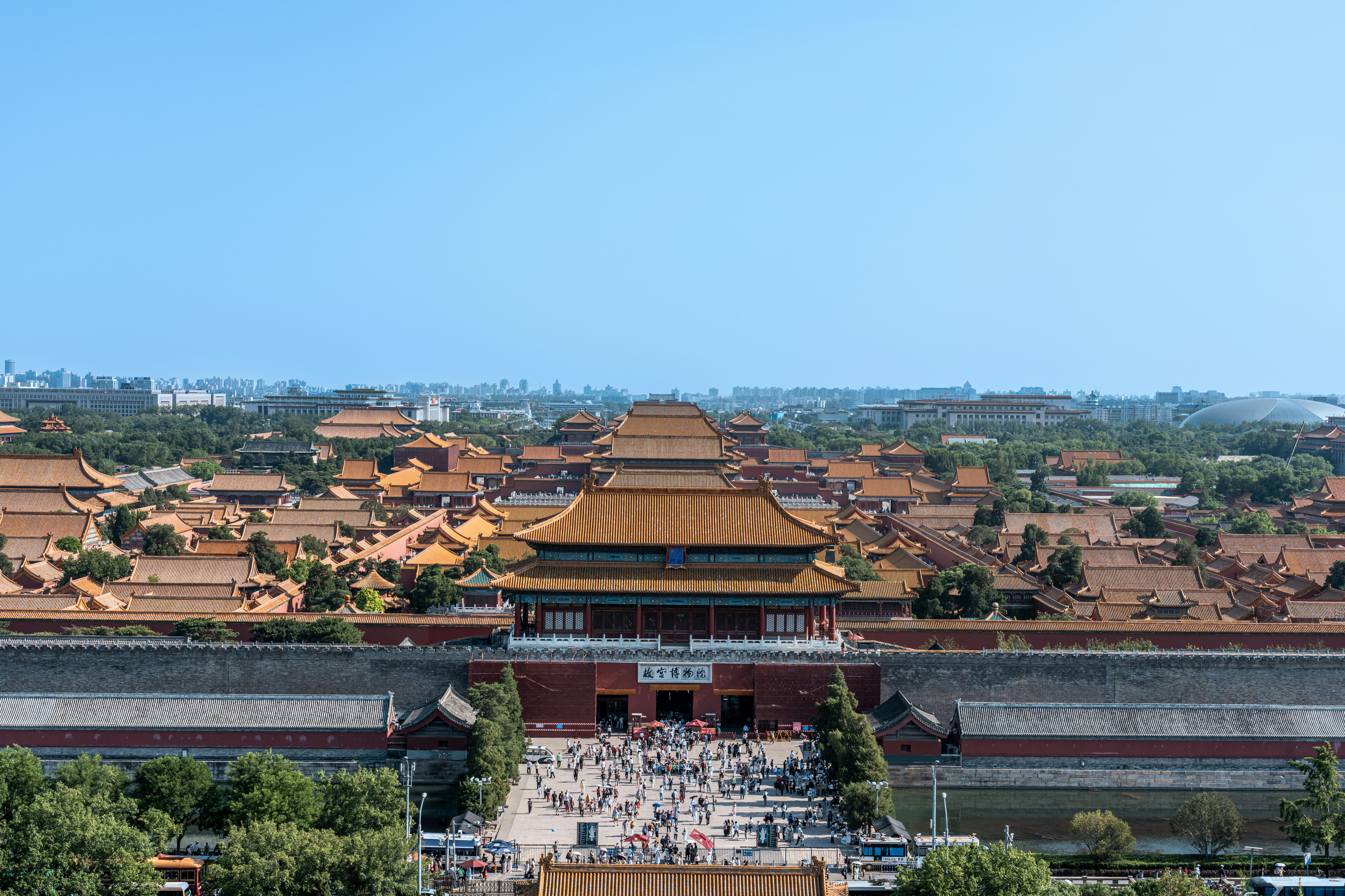 Aerial view of the Forbidden City showcasing its iconic rooftops and sprawling courtyards, bustling with visitors. The clear blue sky highlights the grandeur of this historical site.