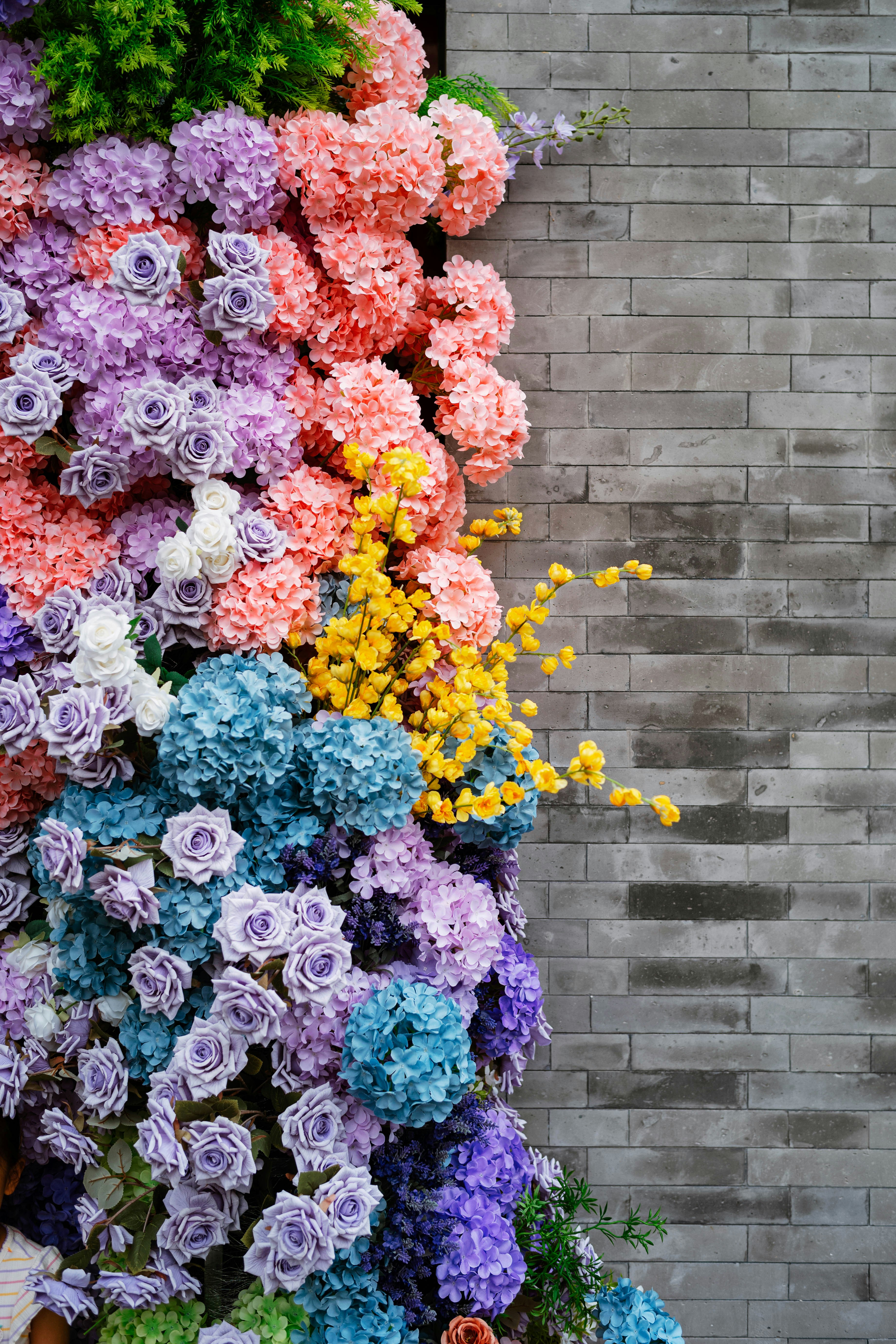 A vibrant arrangement of artificial flowers in various colors, cascading down a textured gray wall. The composition showcases a mix of hydrangeas, roses, and orchids.
