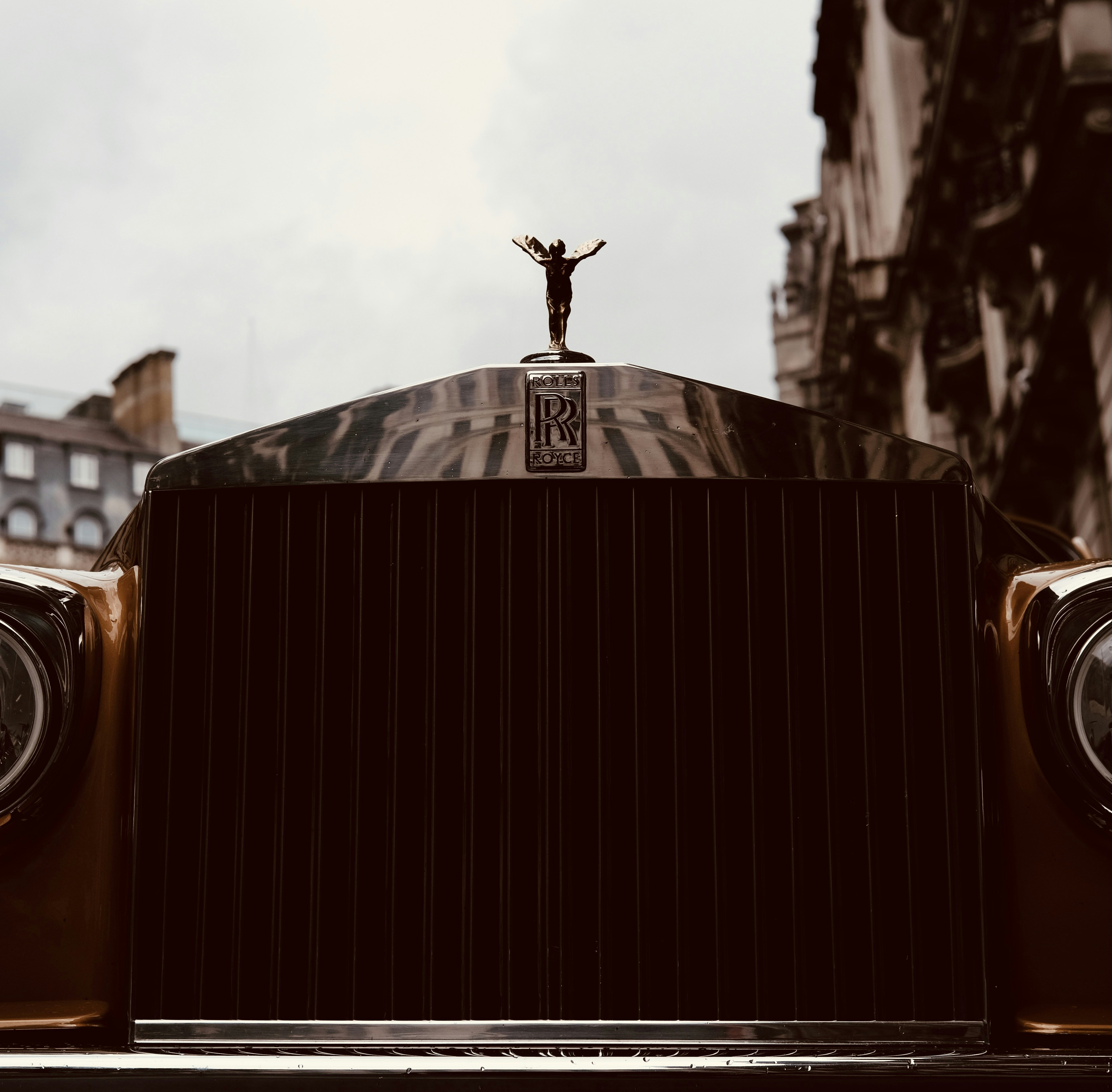 Nahaufnahme eines Rolls-Royce-Kühlergrills mit dem ikonischen Emblem 'Spirit of Ecstasy', umgeben von klassischer Architektur – Symbol für Prestige und Luxus | A rolls royce car stands proudly.