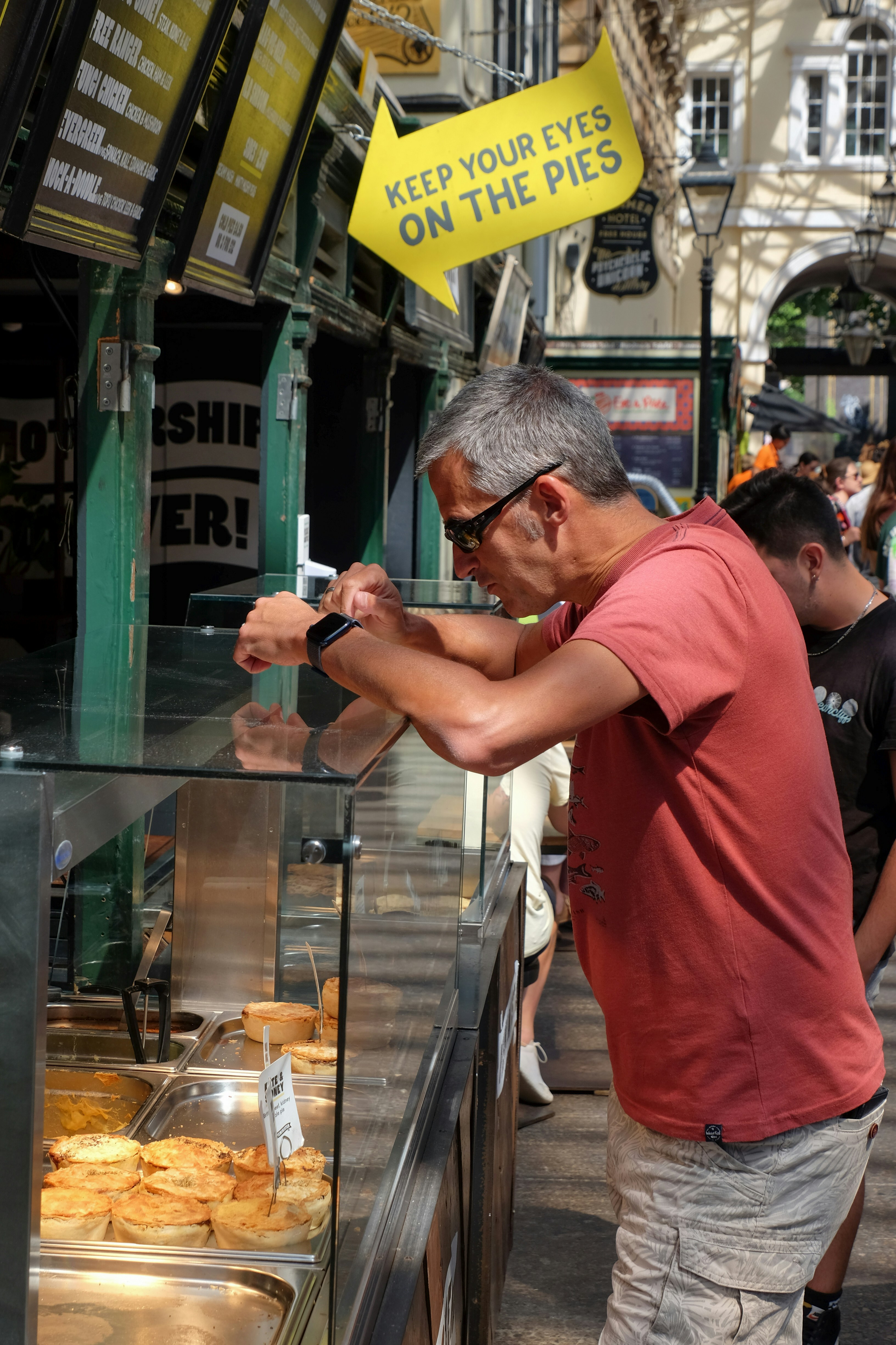 A man examines savory pies at a bustling market stall, with a playful sign urging customers to keep their eyes on the pies.
