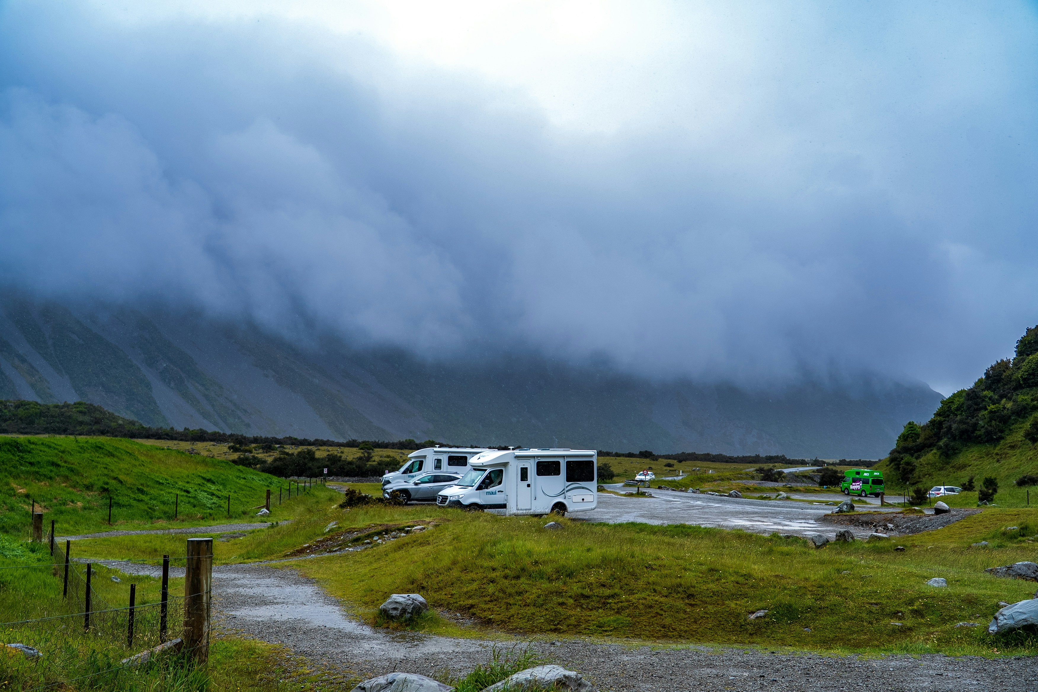 Camping area nestled at the base of towering mountains, shrouded in mist and clouds. Several camper vans are visible in the foreground.