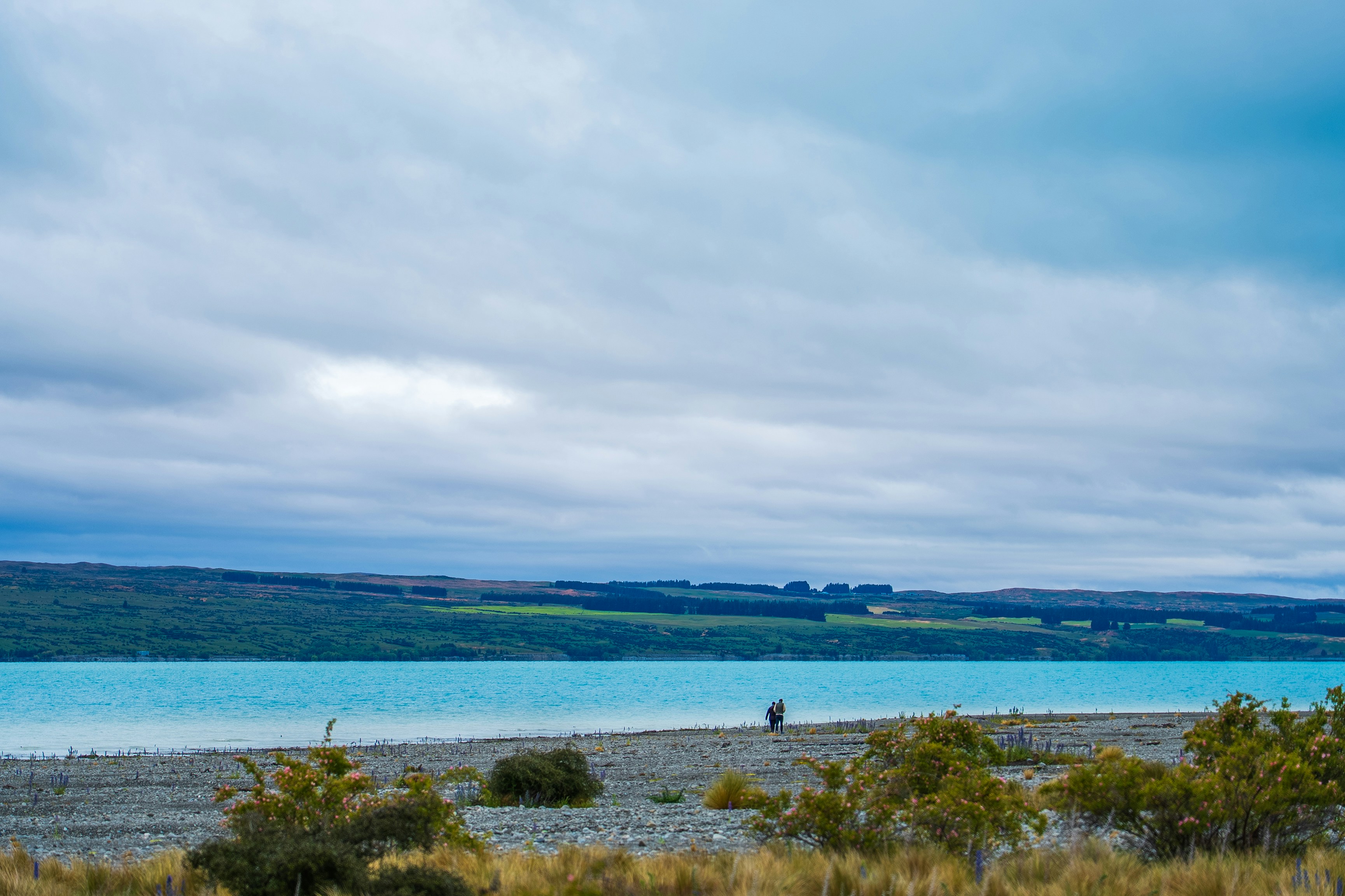 Two figures walking along a pebbled shore under a moody sky, with turquoise waters reflecting the landscape beyond.