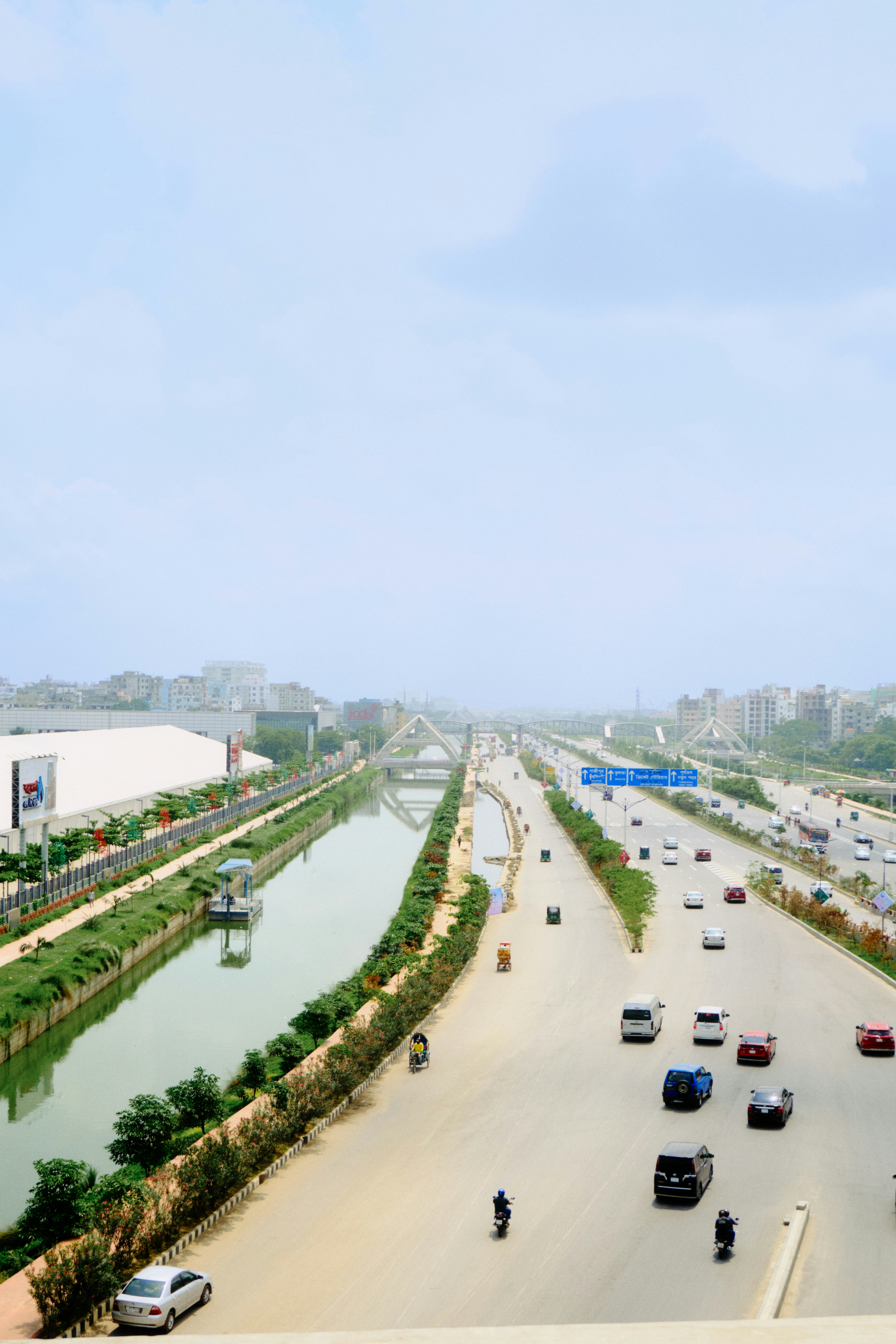 A panoramic view of a bustling urban landscape featuring a winding road alongside a tranquil canal, framed by modern buildings and greenery.