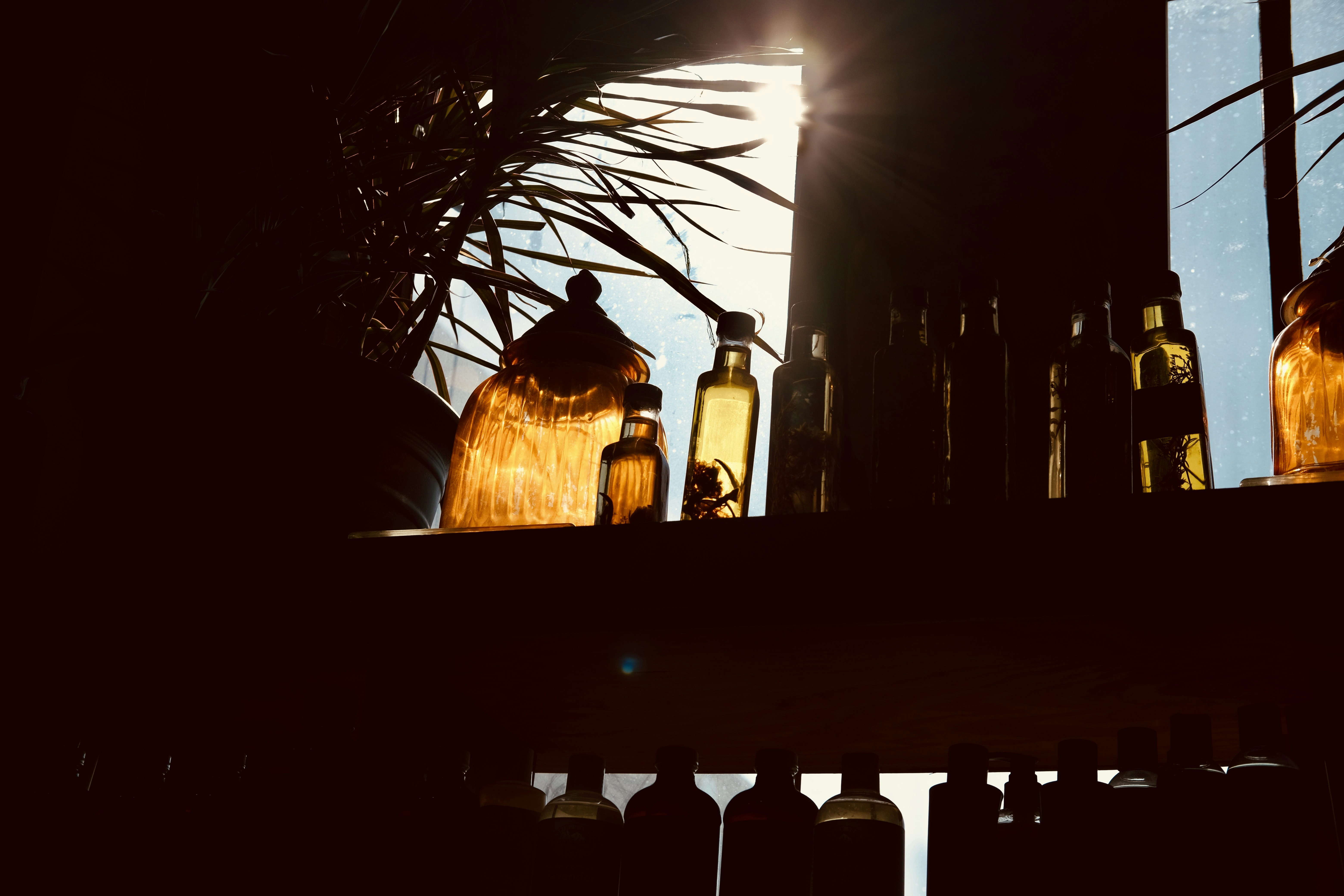 Sonnenlicht trifft auf alte Glasflaschen, gefüllt mit Öl, Kräutern und Erinnerungen. Dieses Bild fängt nicht nur ein Regal ein – sondern das, was zwischen Licht und Dunkelheit passiert. Eine stille Ode an Einfachheit, Handwerk und langsame Räume. | Bottles lined up in front of bright sunlight.