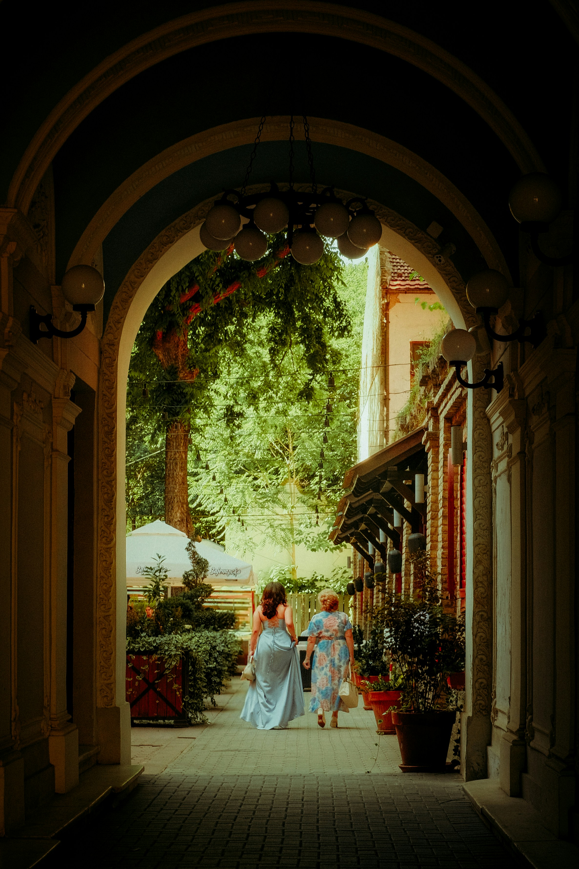 Two women walk through a charming archway adorned with greenery, leading into a vibrant courtyard filled with plants and rustic decor.