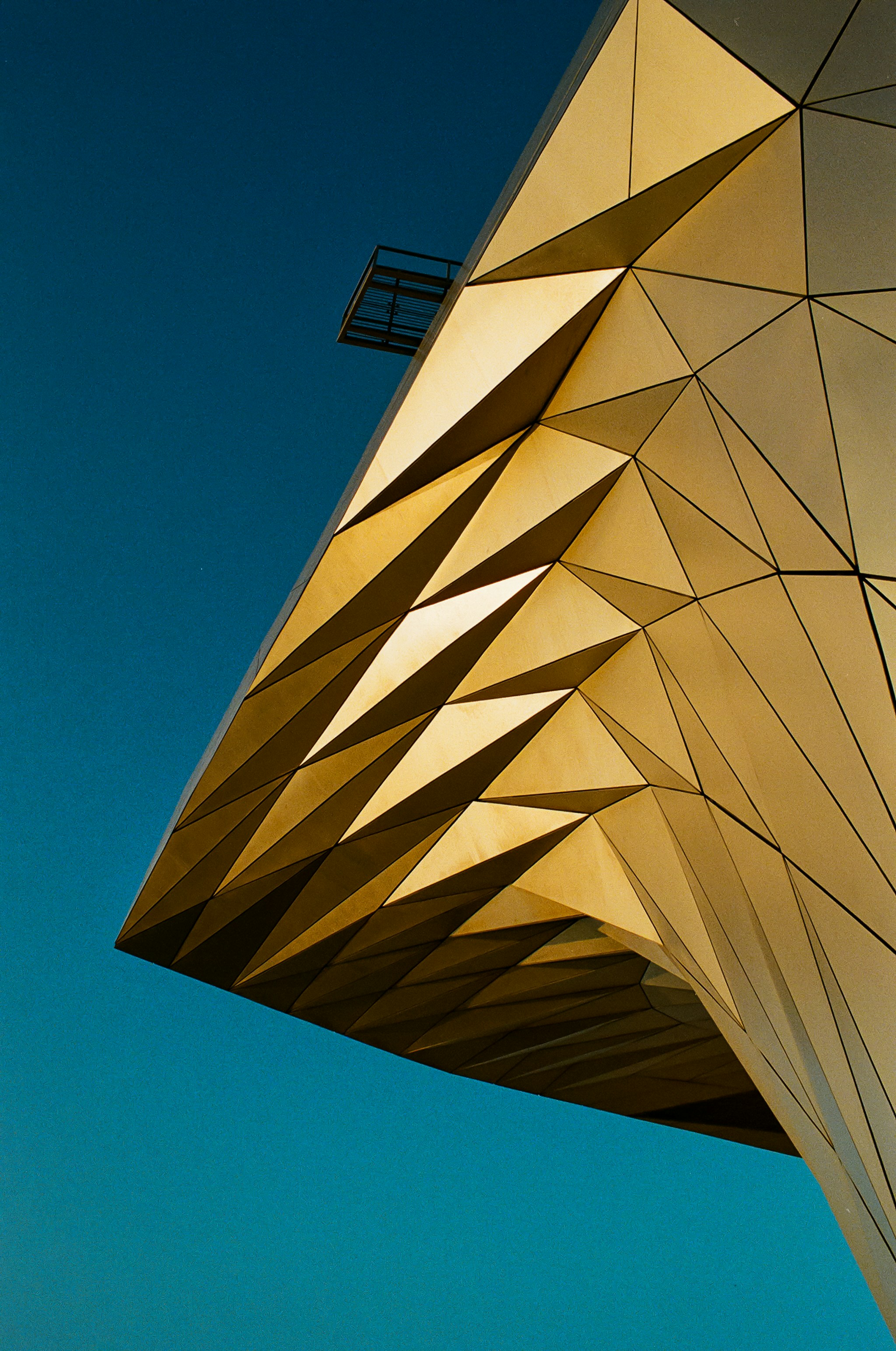 Angular metallic structure reflecting warm light against a deep blue sky. The intricate patterns create a dynamic visual experience.