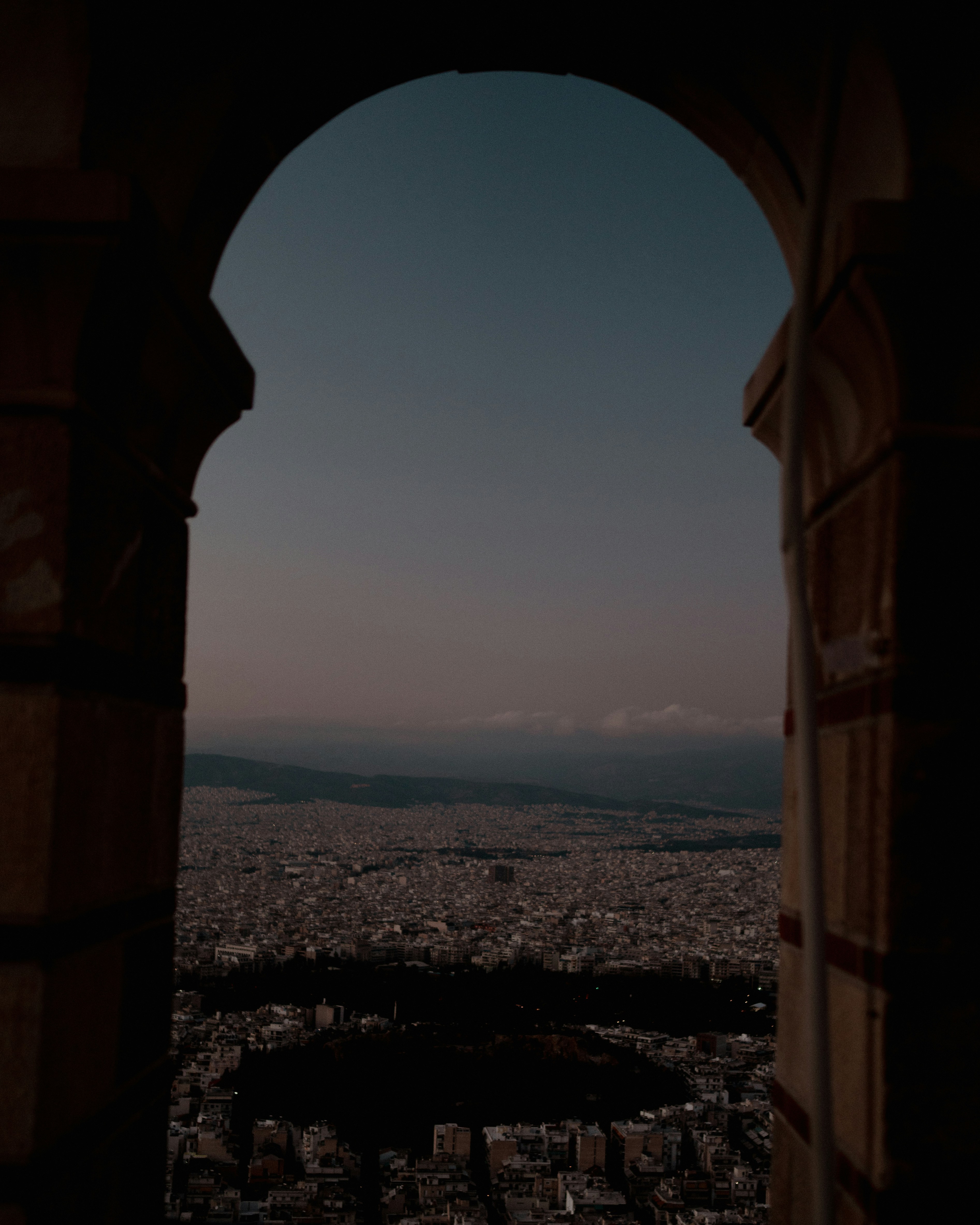 View from an ancient archway overlooking a sprawling cityscape at twilight. The soft gradient of the sky adds depth to the scene.