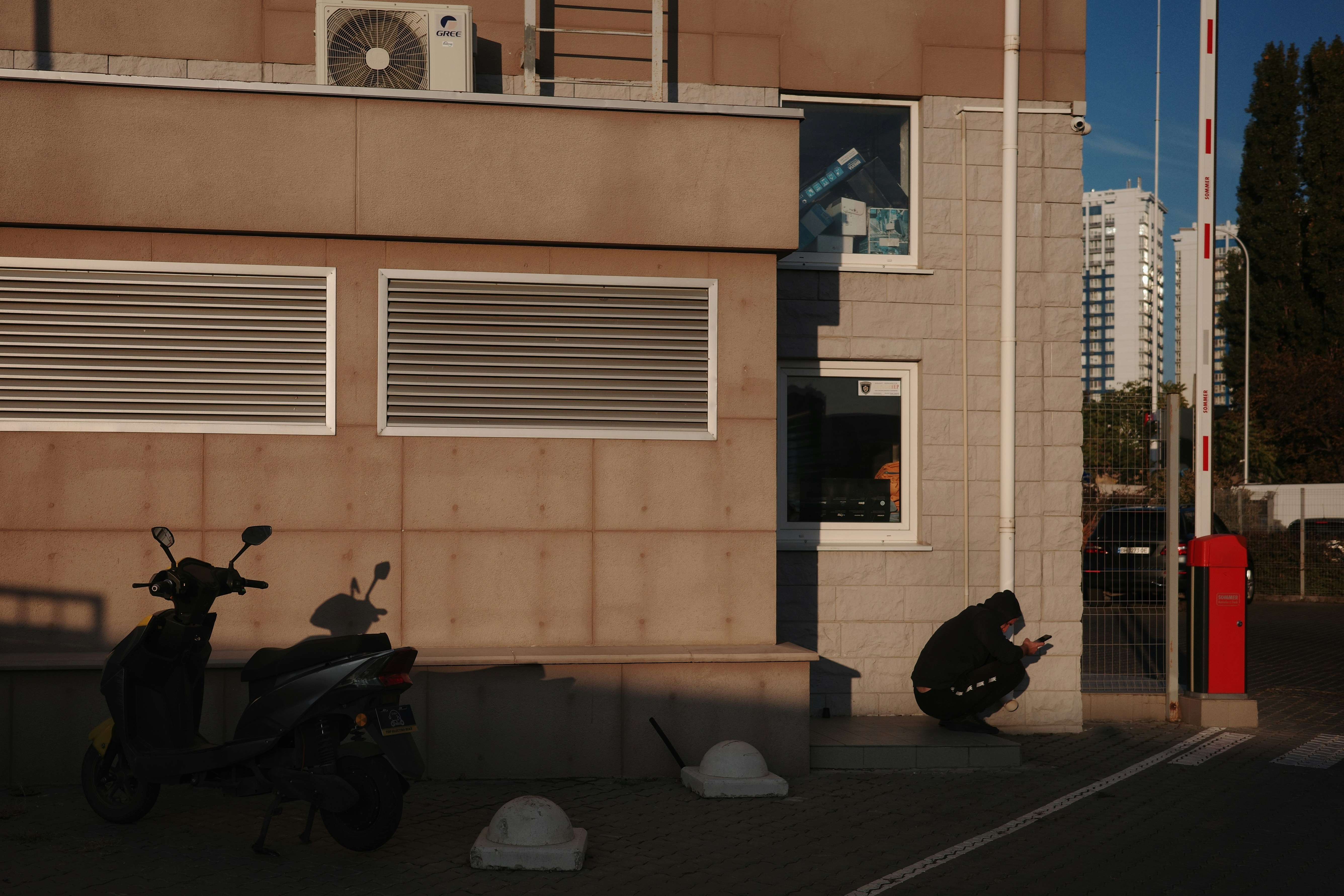 A lone figure crouches beside a building, absorbed in their phone, while a scooter rests nearby. The interplay of light and shadow creates a contemplative atmosphere.