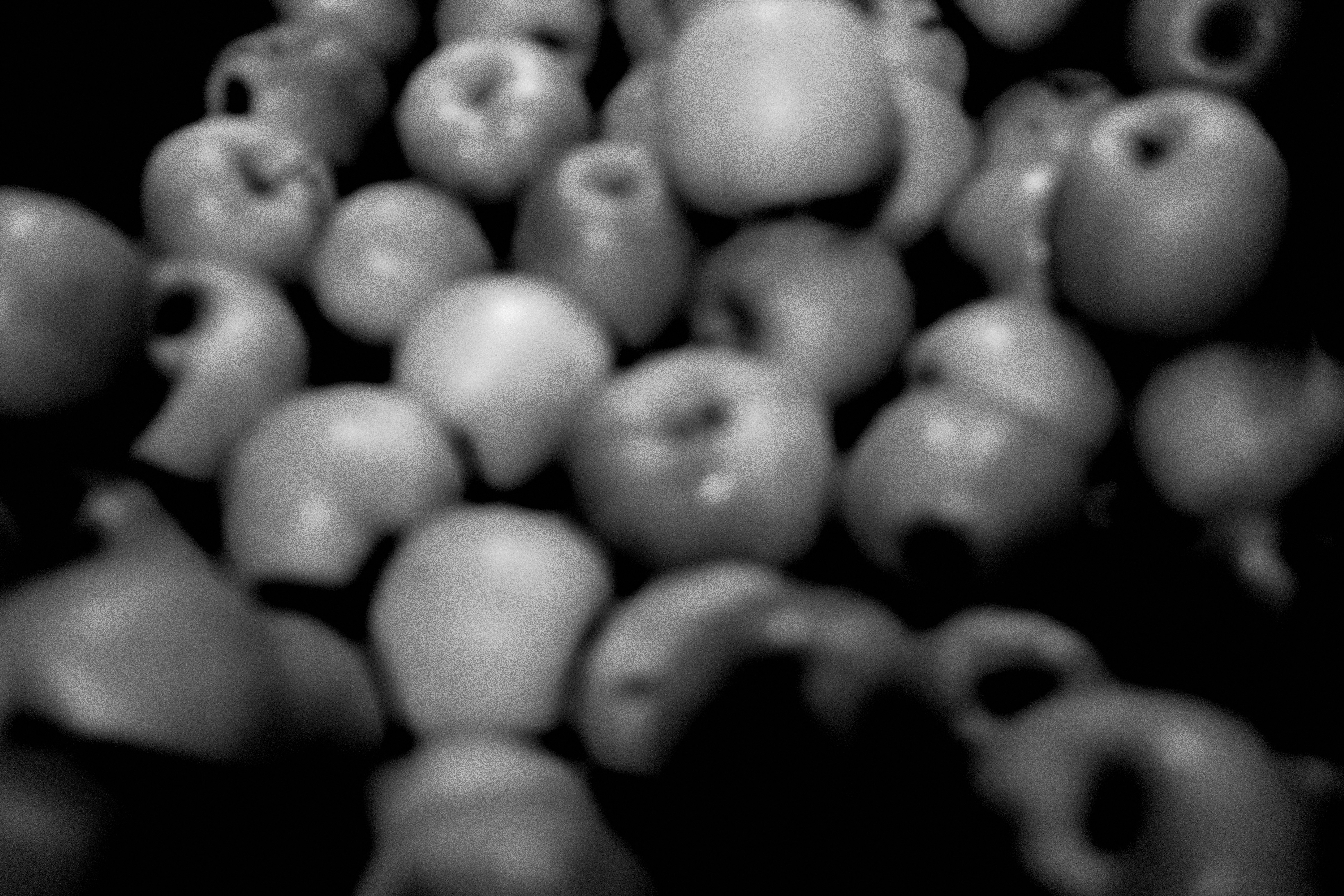 A high-angle, black and white shot captures a scattered collection of small, apple-shaped objects, possibly beads or decorative elements, against a dark background. Many of the objects appear to have a small hole, suggesting they are designed to be strung or attached. The image is intentionally out of focus, creating a soft, dreamlike, and somewhat abstract quality. | Many apples are scattered in this blurry image.