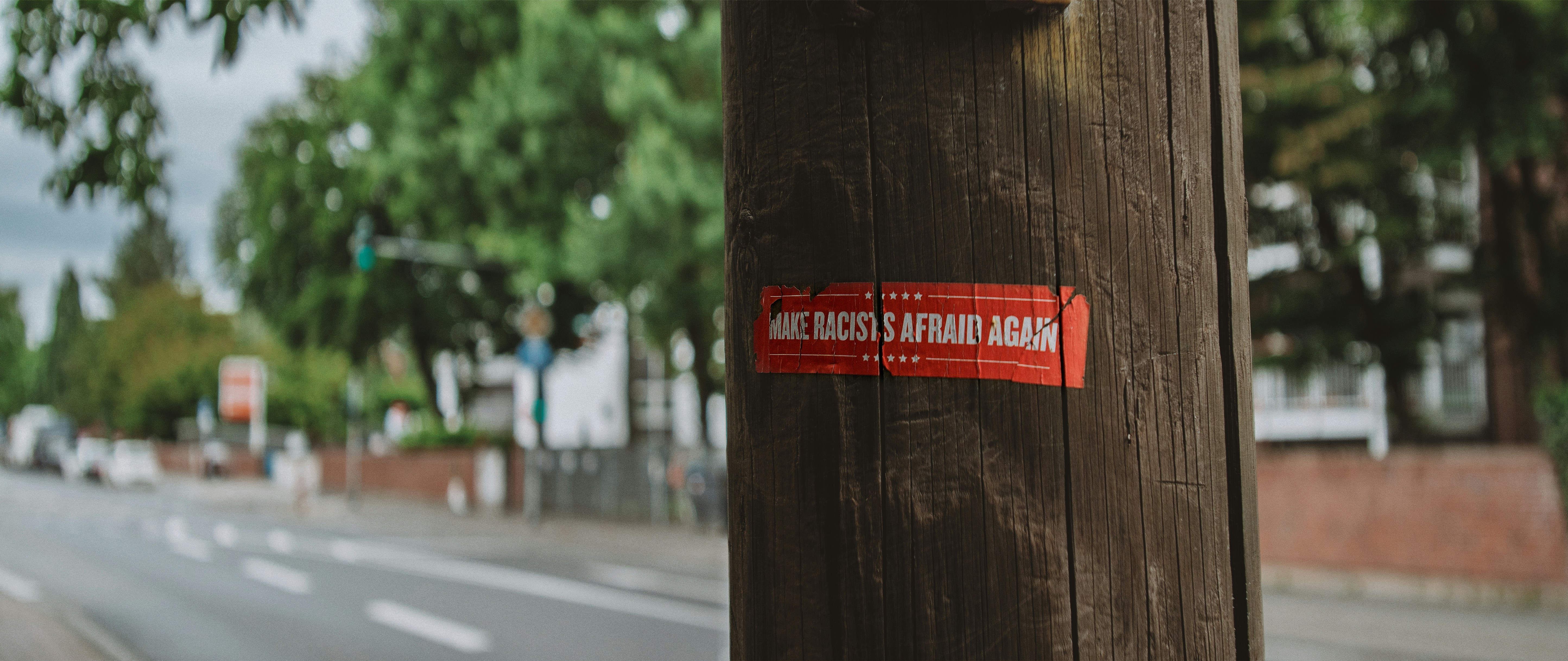A striking red sticker on a wooden post conveys a powerful message in an urban setting. The background features a blurred street scene with greenery.