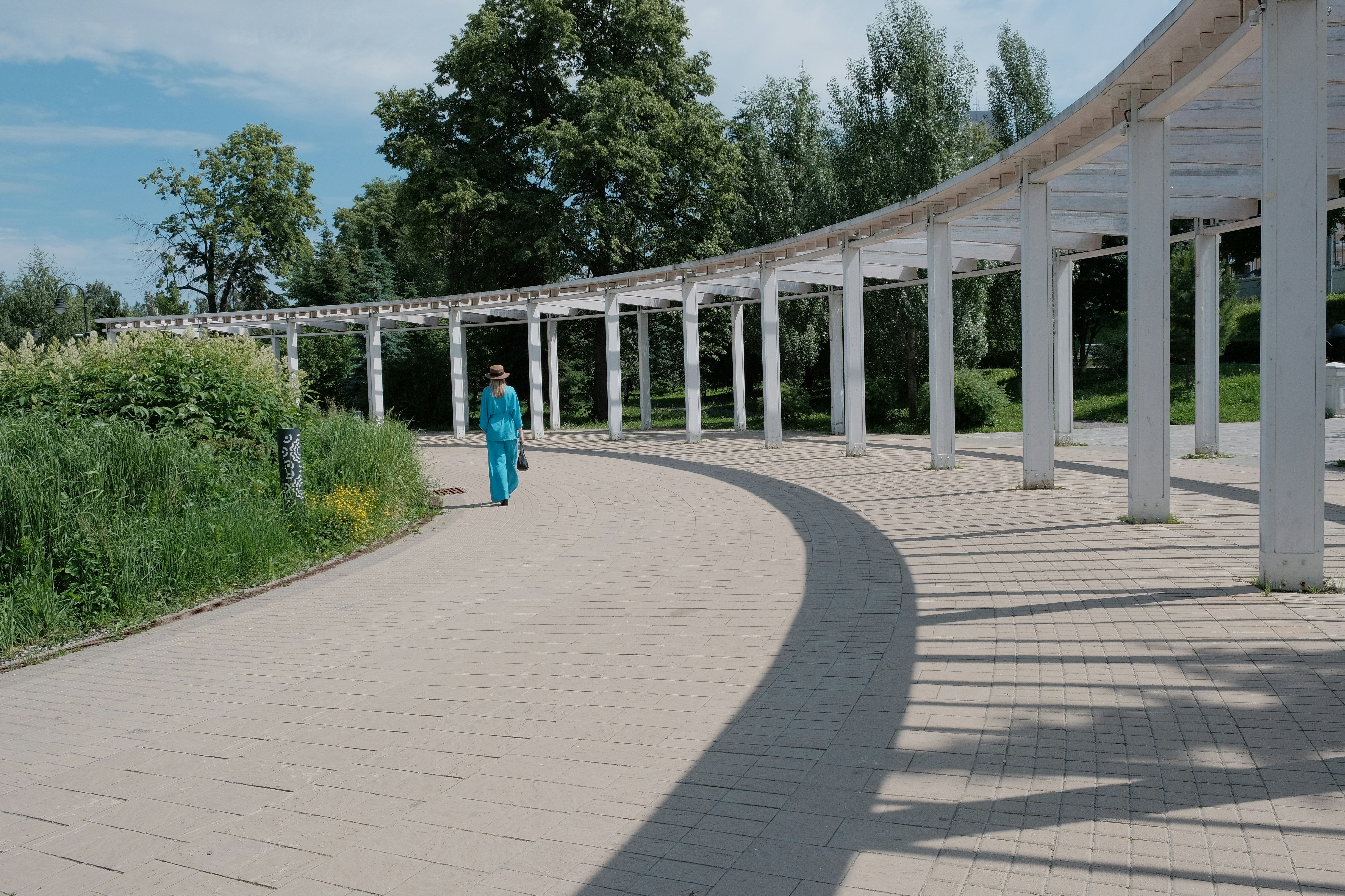 A person in a bright blue outfit walks along a paved path under a white pergola, surrounded by greenery and trees on a sunny day. | A person walks along a curved pathway.