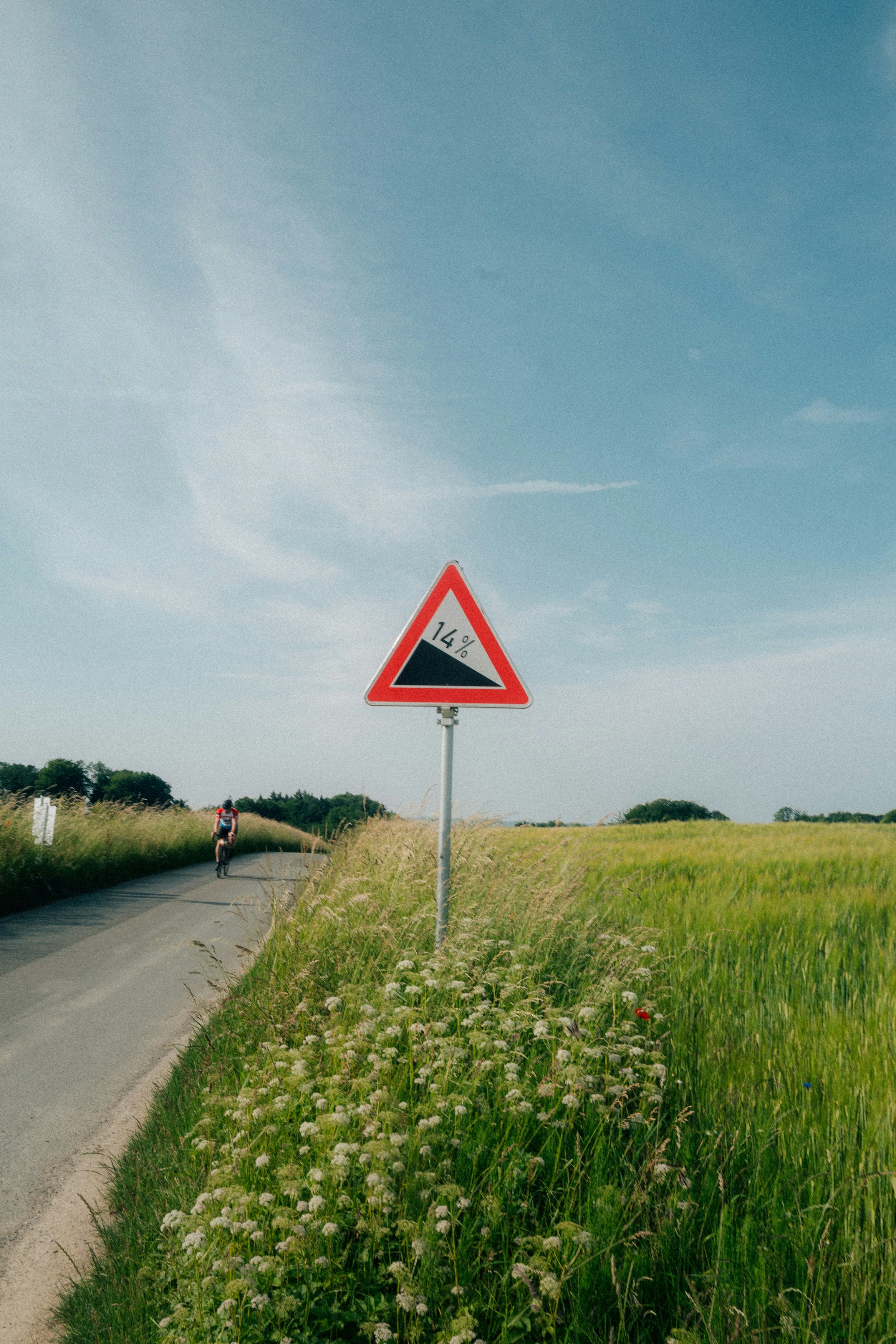 Warning sign indicating a steep gradient on a country road, with a cyclist approaching amidst lush greenery.