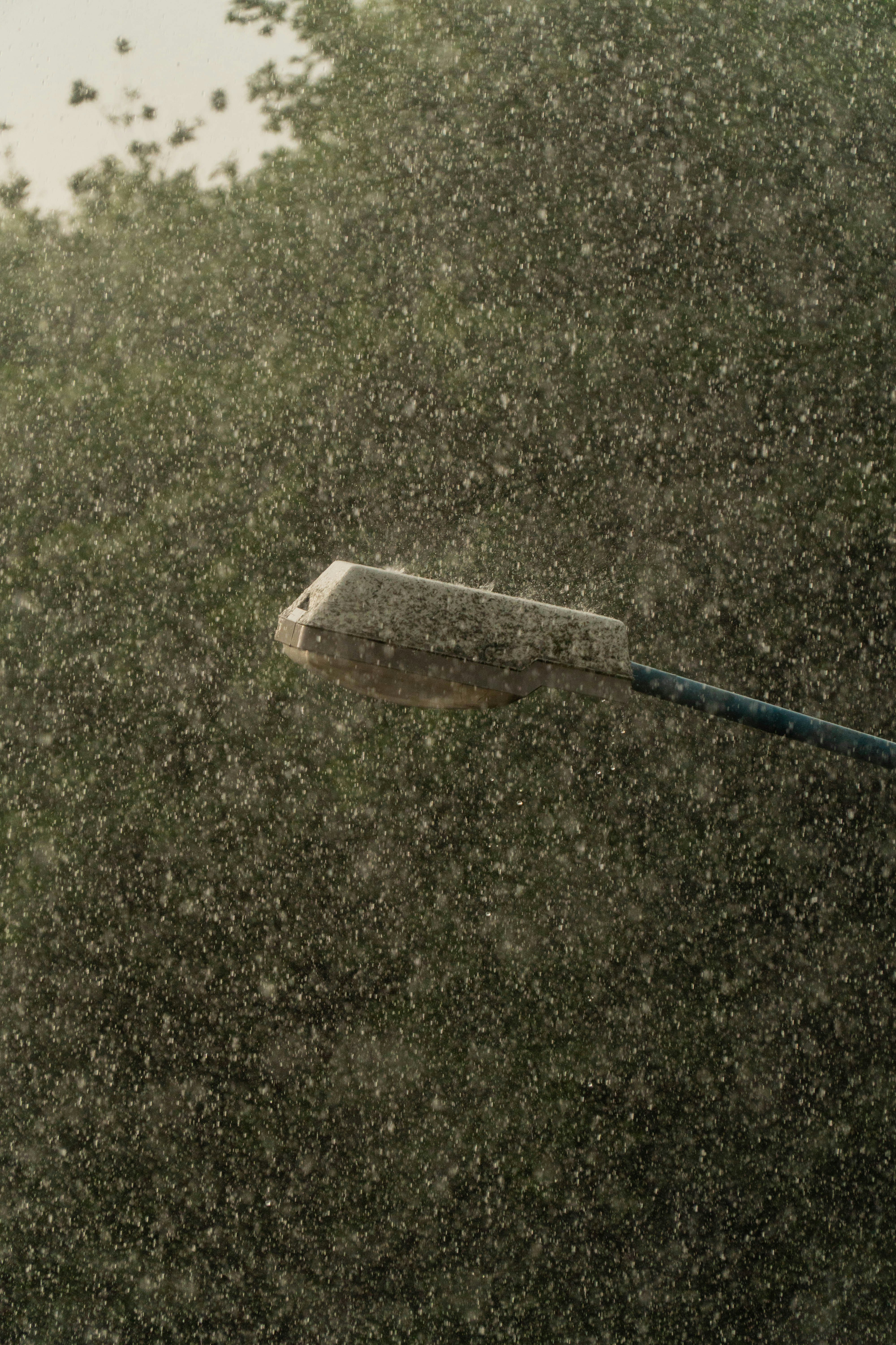 A weathered streetlight stands amidst a cascade of raindrops, creating a serene atmosphere in an urban landscape. The blurred background enhances the focus on the light's texture.