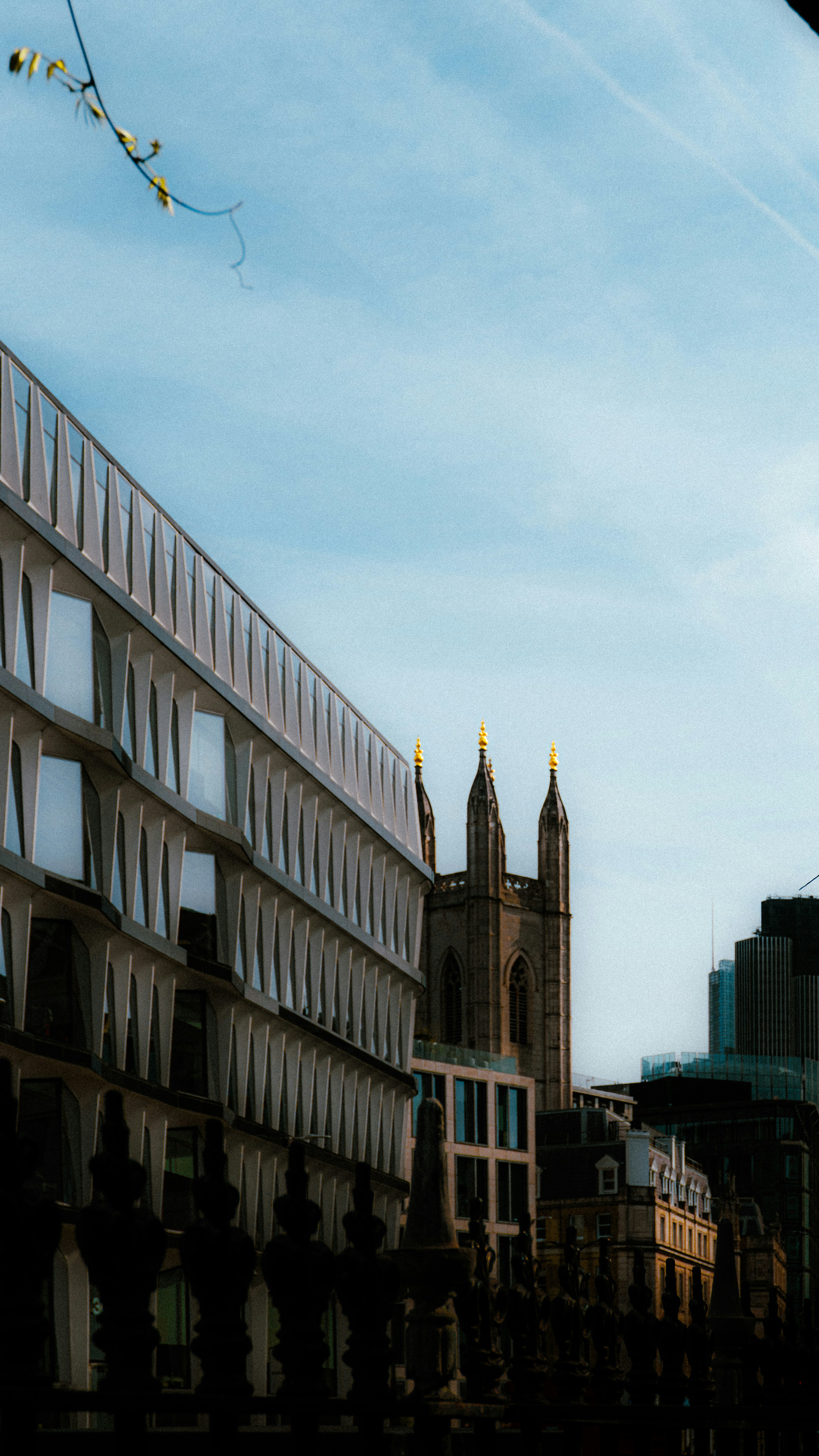 A juxtaposition of old and new architecture in London, where a modern geometric facade meets the gothic towers of a historic church. The golden spires glint subtly under a clear blue sky, capturing the layered character of the cityscape. | Modern buildings contrast with an old church's spires.