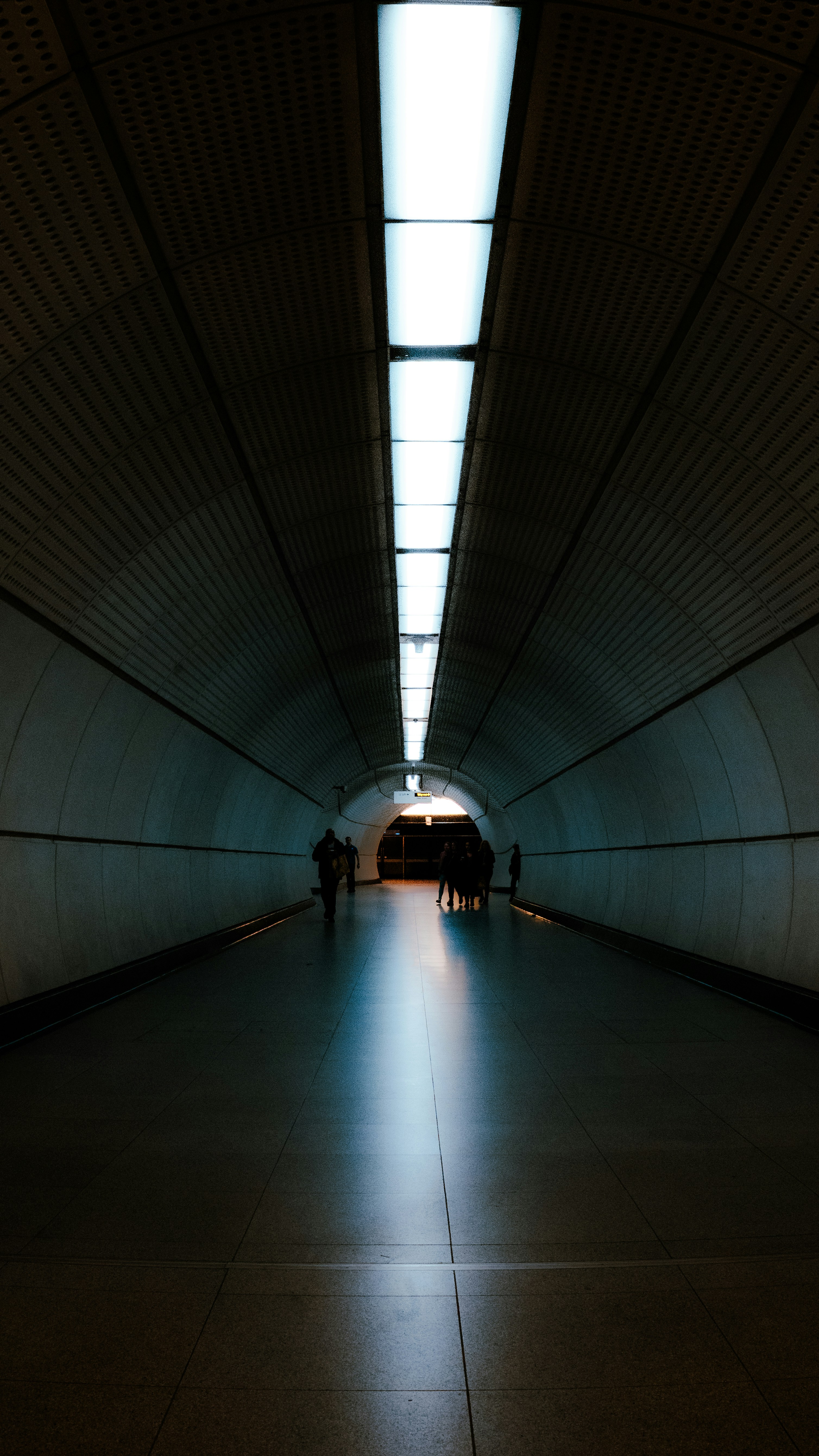 Dimly lit underground tunnel with a long perspective, featuring silhouetted figures walking towards a bright exit. 