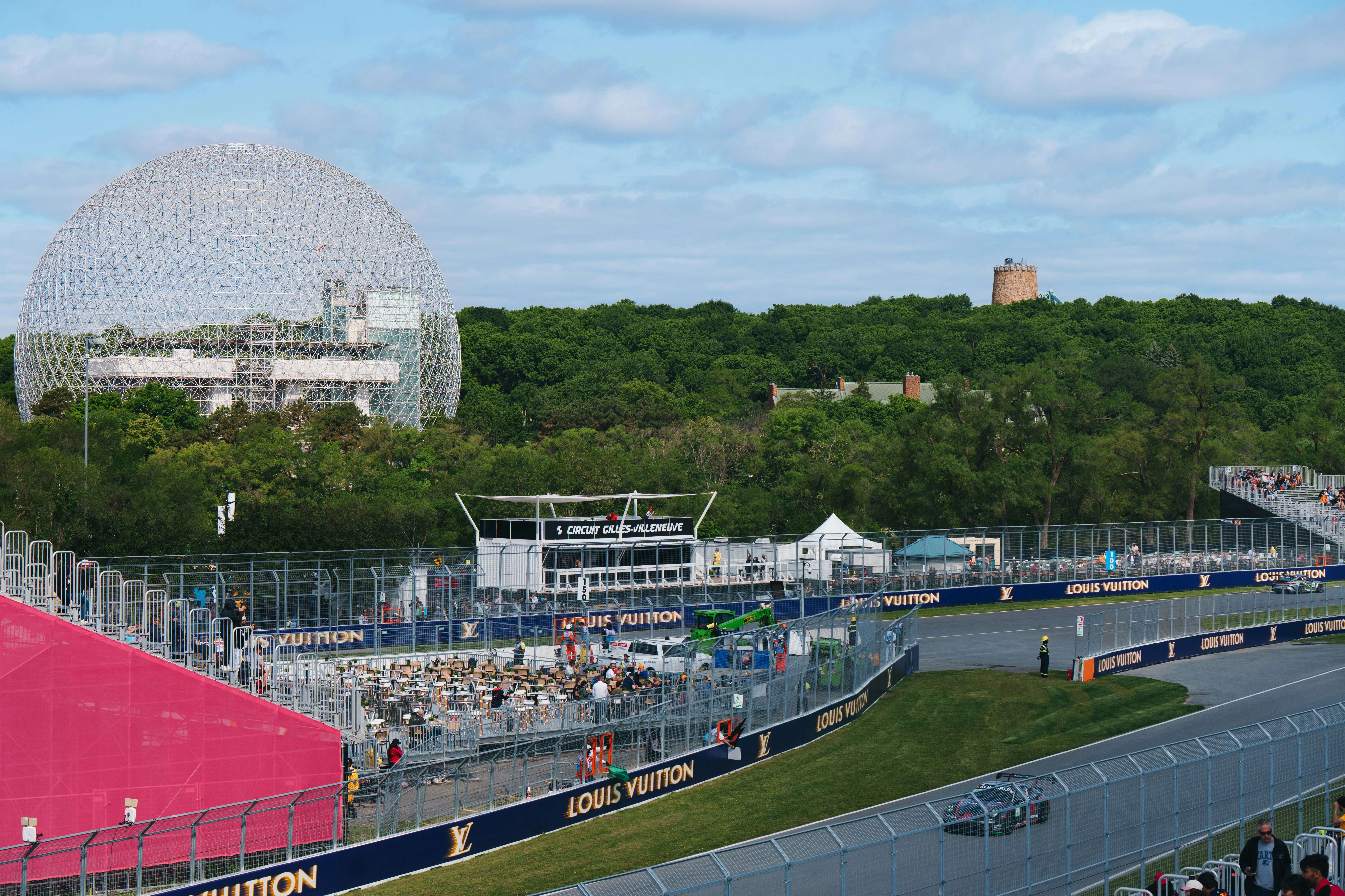 Race cars navigate a circuit surrounded by lush greenery, with an iconic geodesic dome in the background. The scene captures the dynamic energy of motorsport against a tranquil landscape.