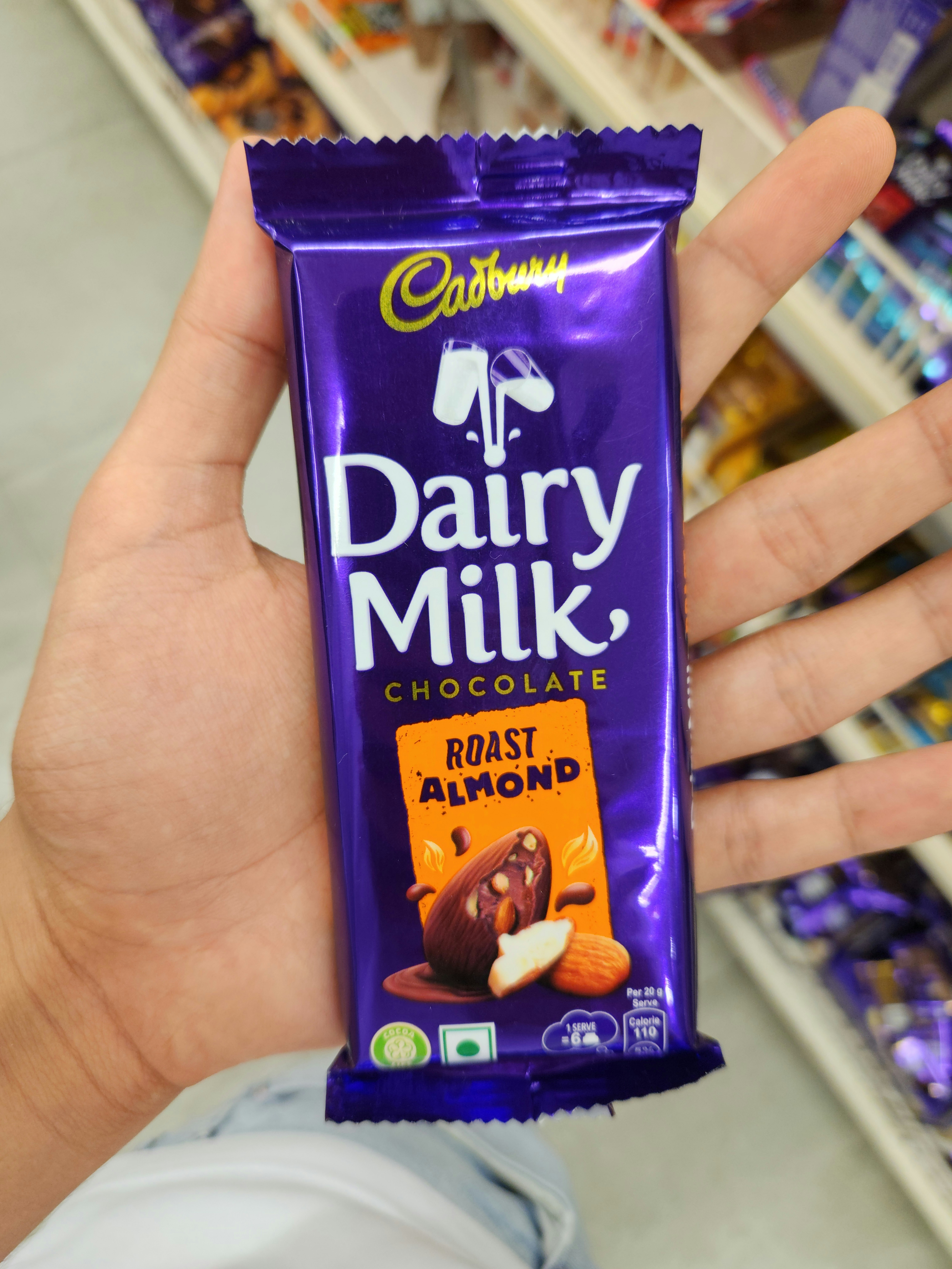 The image shows a Cadbury Dairy Milk Roast Almond chocolate bar, a popular Indian variant of the classic milk chocolate. The packaging features Cadbury’s signature purple color, with visual cues of roasted almonds and rich chocolate. Known for its creamy texture and crunchy almond bits, this bar is a favorite among chocolate lovers in India. The photo captures both the branding and the indulgent appeal of this widely enjoyed confectionery product.