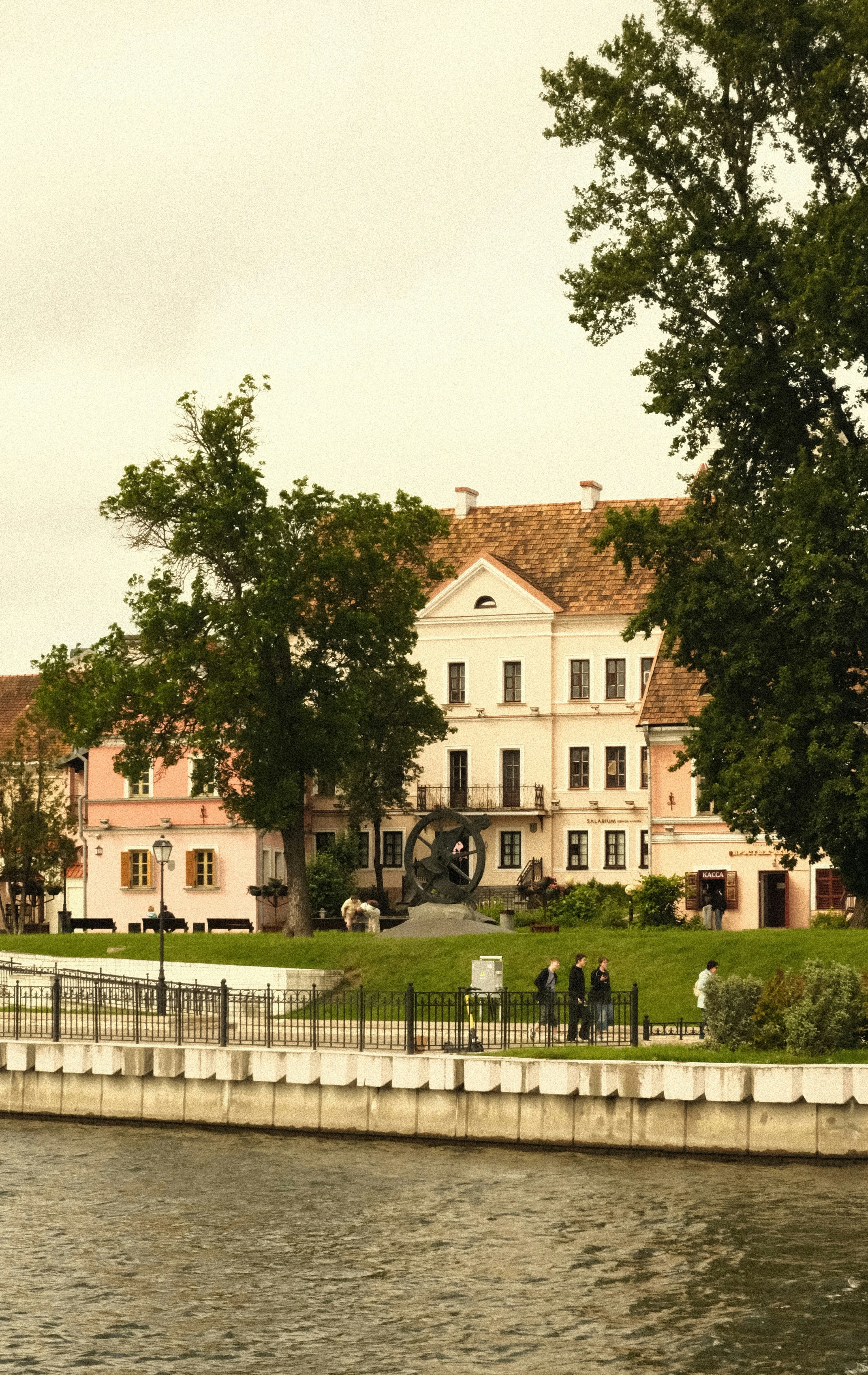 Charming riverside scene featuring a historical building with lush greenery and a sculpture, inviting visitors to explore the serene environment.