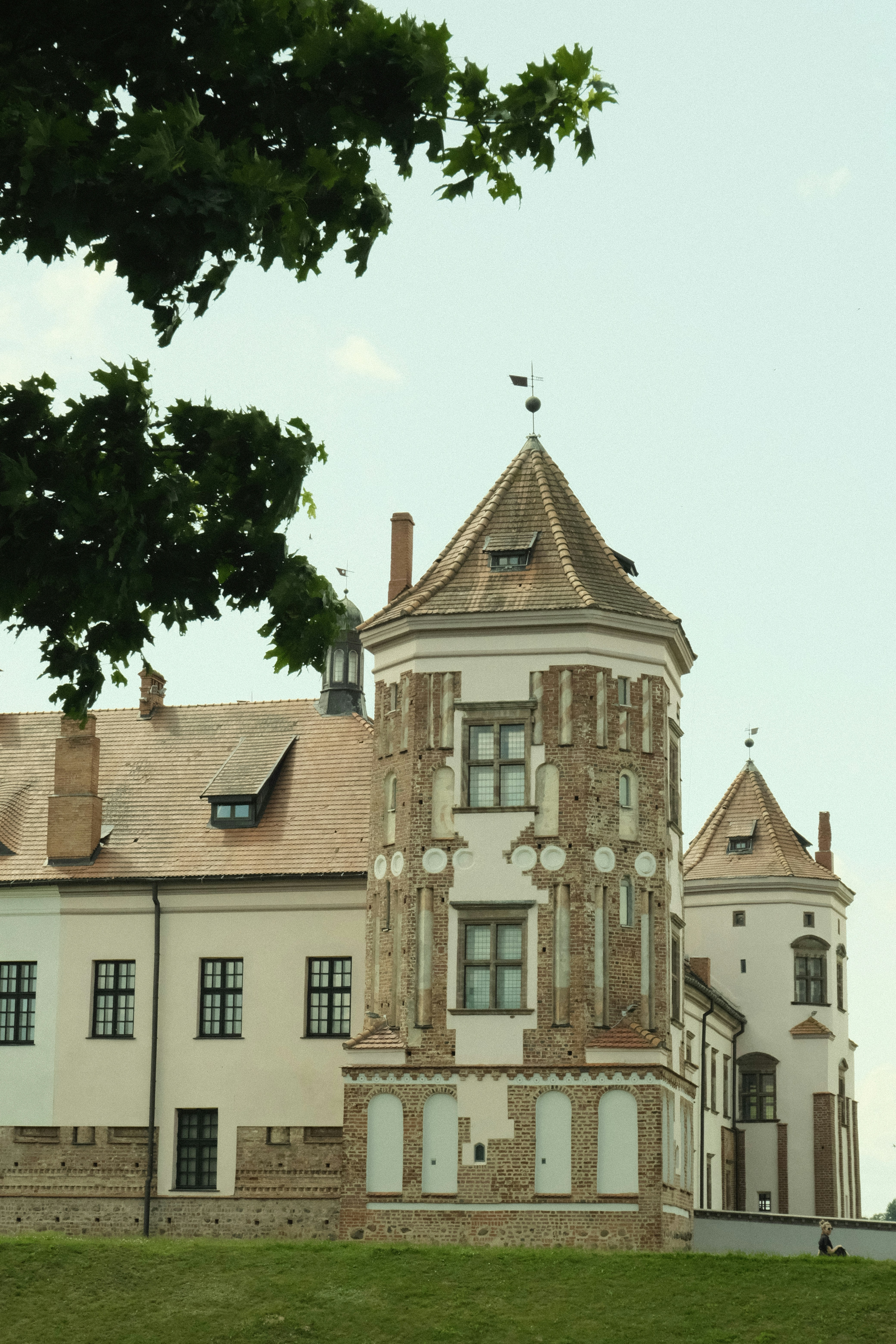 Historic tower with distinctive brickwork and roof, standing beside a classic building amidst greenery.