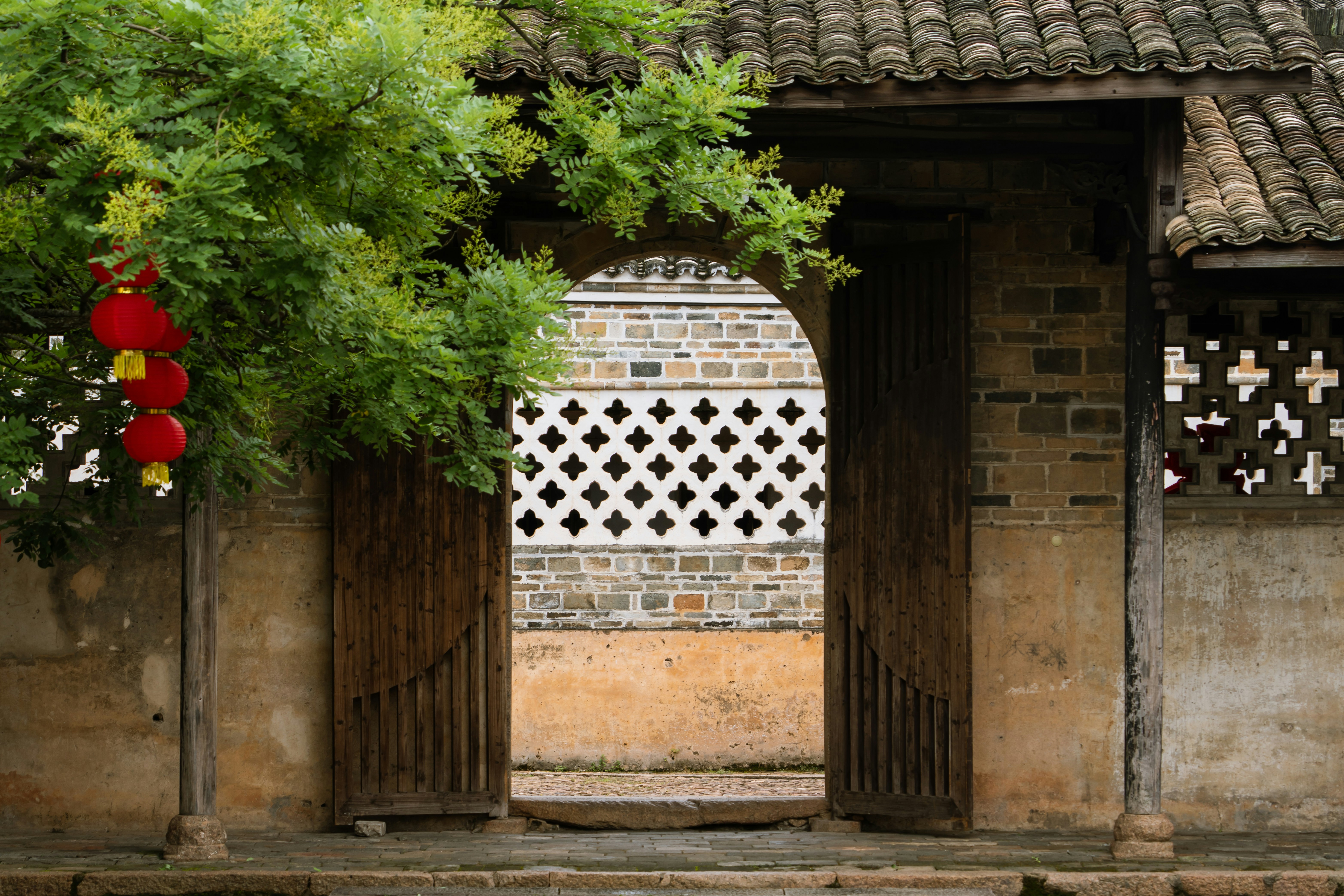Arched wooden doorway framed by lush greenery and traditional lanterns, revealing a patterned wall beyond. The scene evokes a sense of cultural heritage.