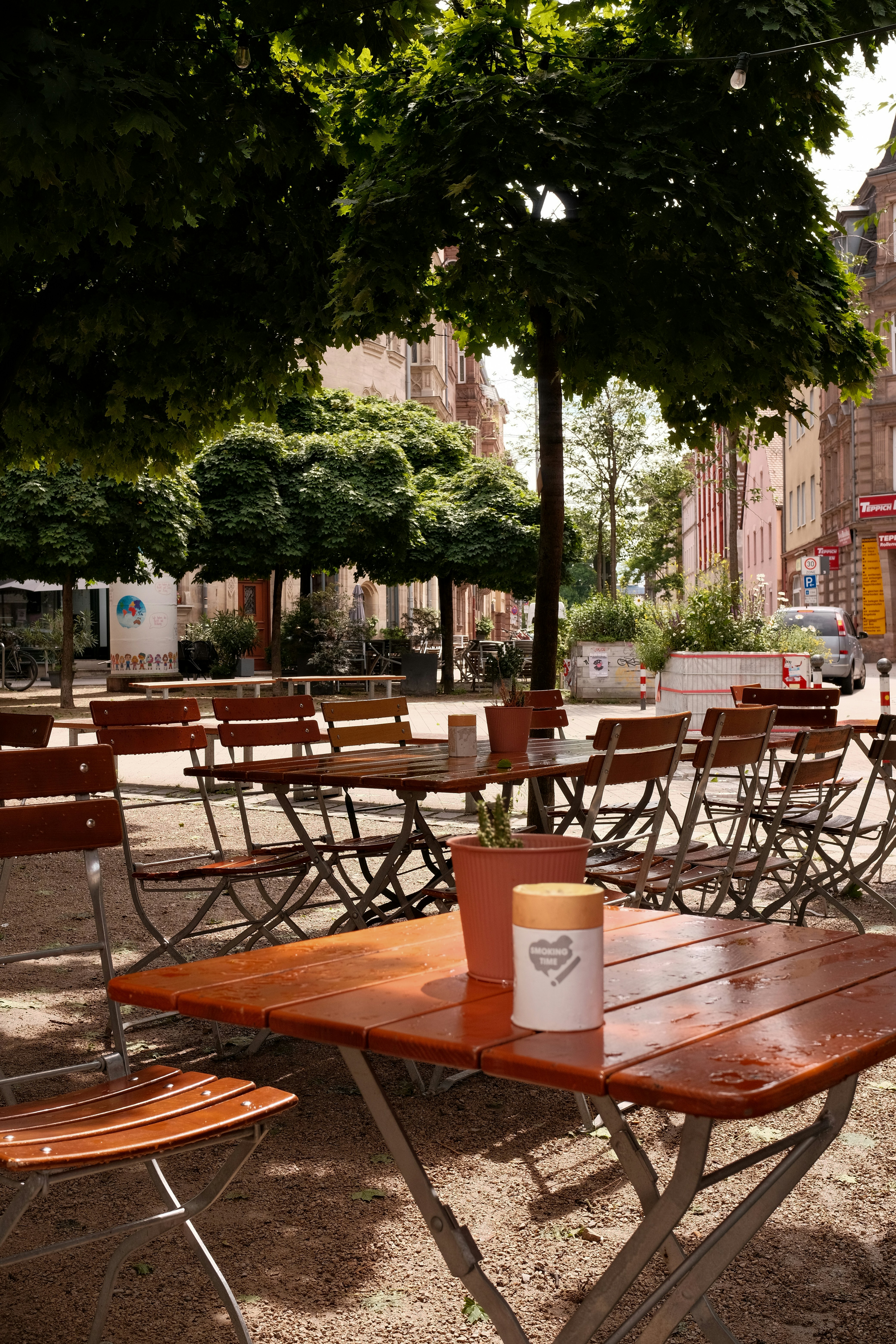 Charming outdoor dining area in a quiet urban plaza surrounded by lush green trees and wooden tables | Outdoor cafe with tables and chairs under the trees.