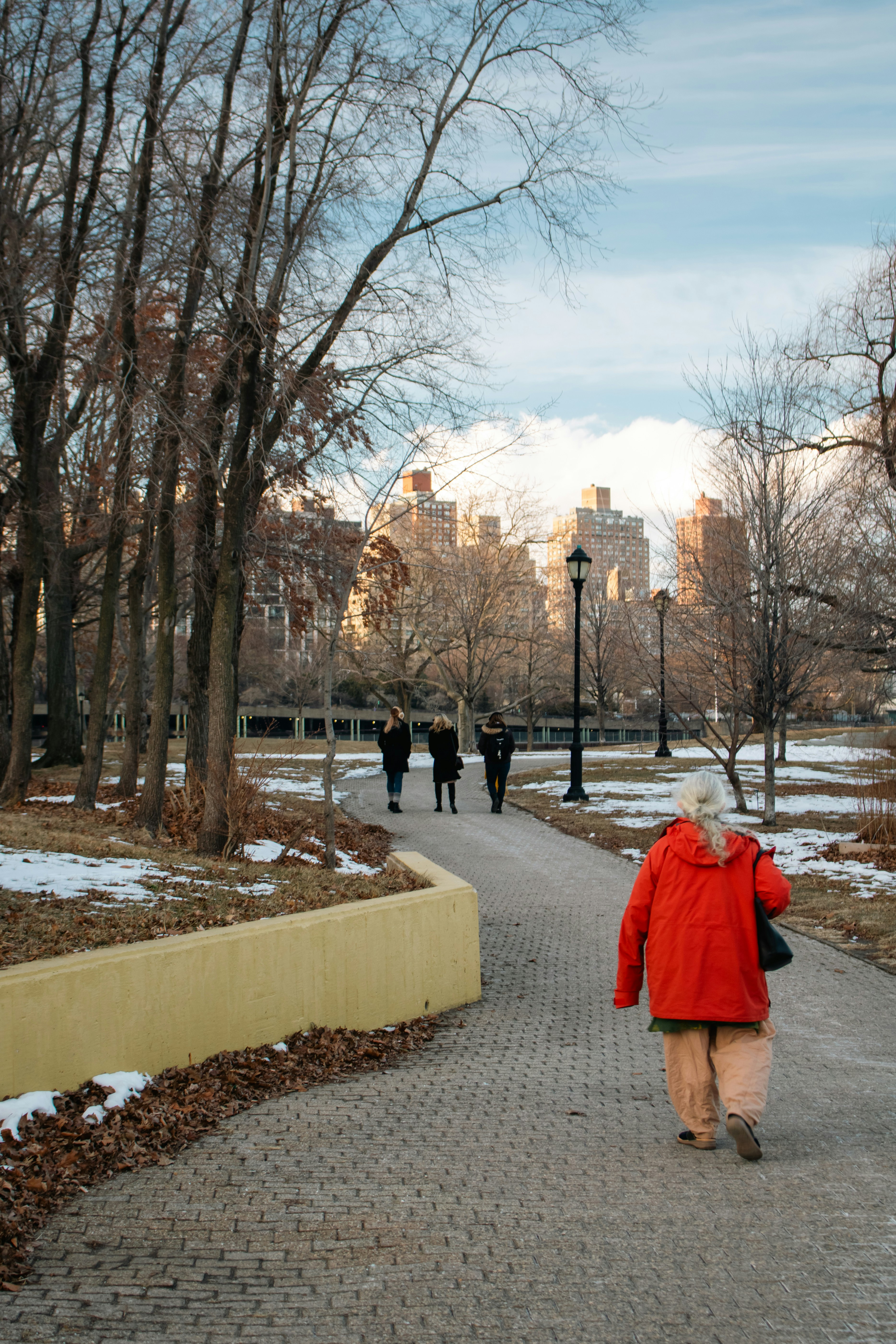 A winter scene on Roosevelt Island, New York City. In the foreground, a person in an orange jacket walks away from the camera. Behind them, three individuals stroll together. The ground is dotted with patches of snow, and the bare trees suggest it's late fall or winter. Low stone walls border the path, and buildings are visible in the distance. The image captures a quiet, cold day in an urban park, blending human activity with the stillness of the season. | A person walks down a path in a park.