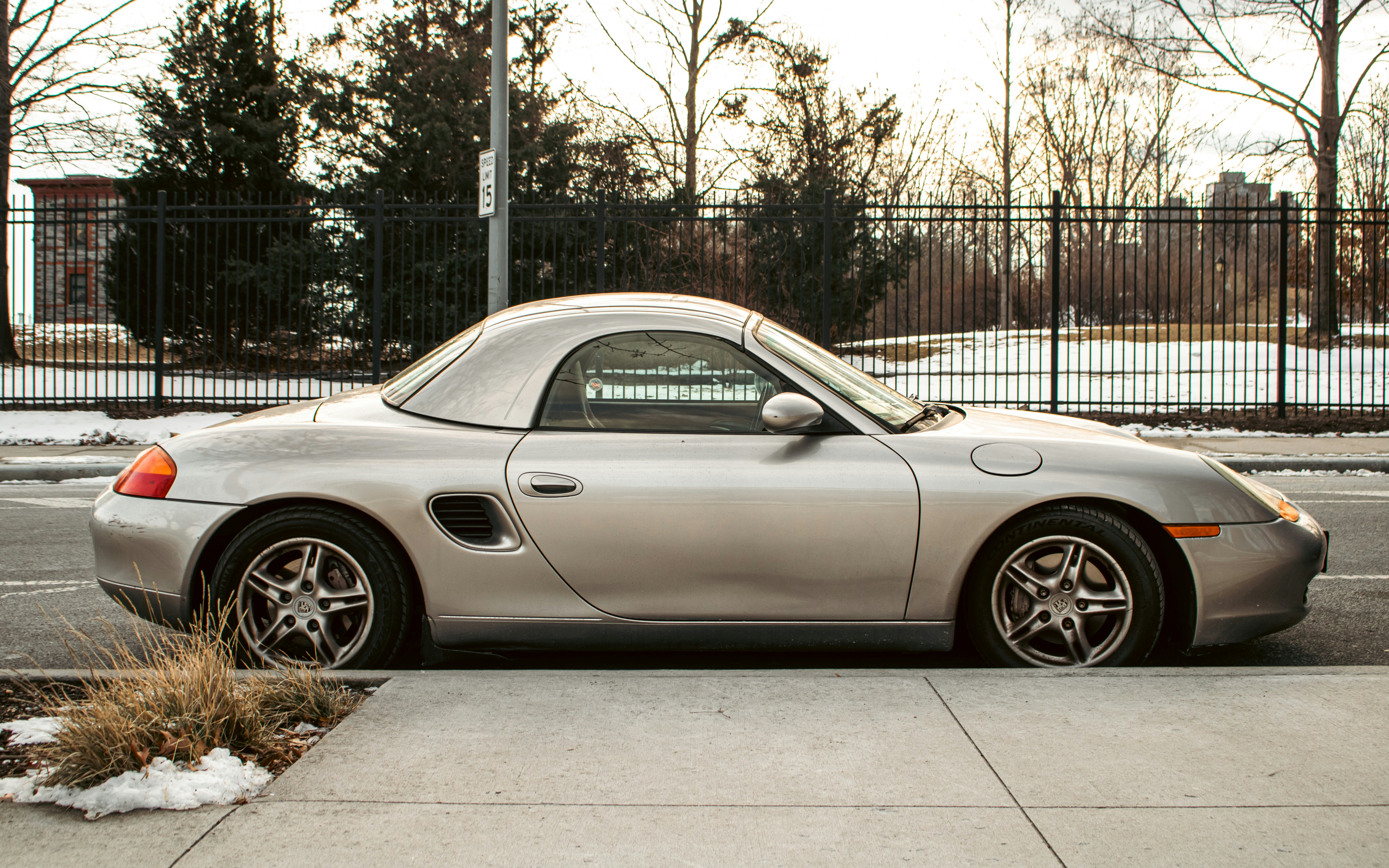 A sleek silver Porsche Boxster is parked along a quiet, empty street on Roosevelt Island, New York City, during a crisp winter day. | A silver convertible car parked on the side of the road.