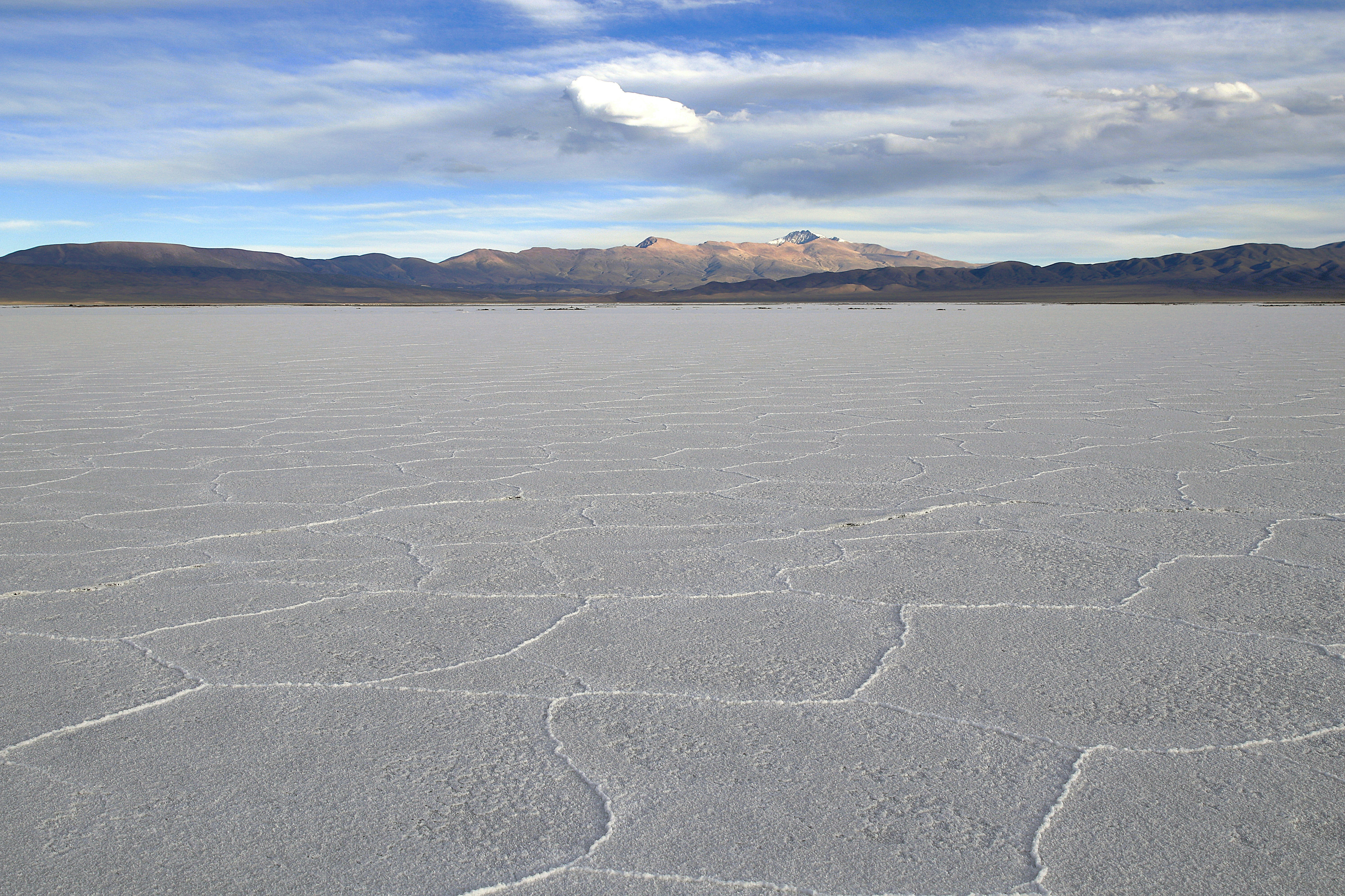 Expansive salt flat stretches under a cloudy sky, with distant mountains providing a serene backdrop. Unique hexagonal patterns of salt create a textured surface.