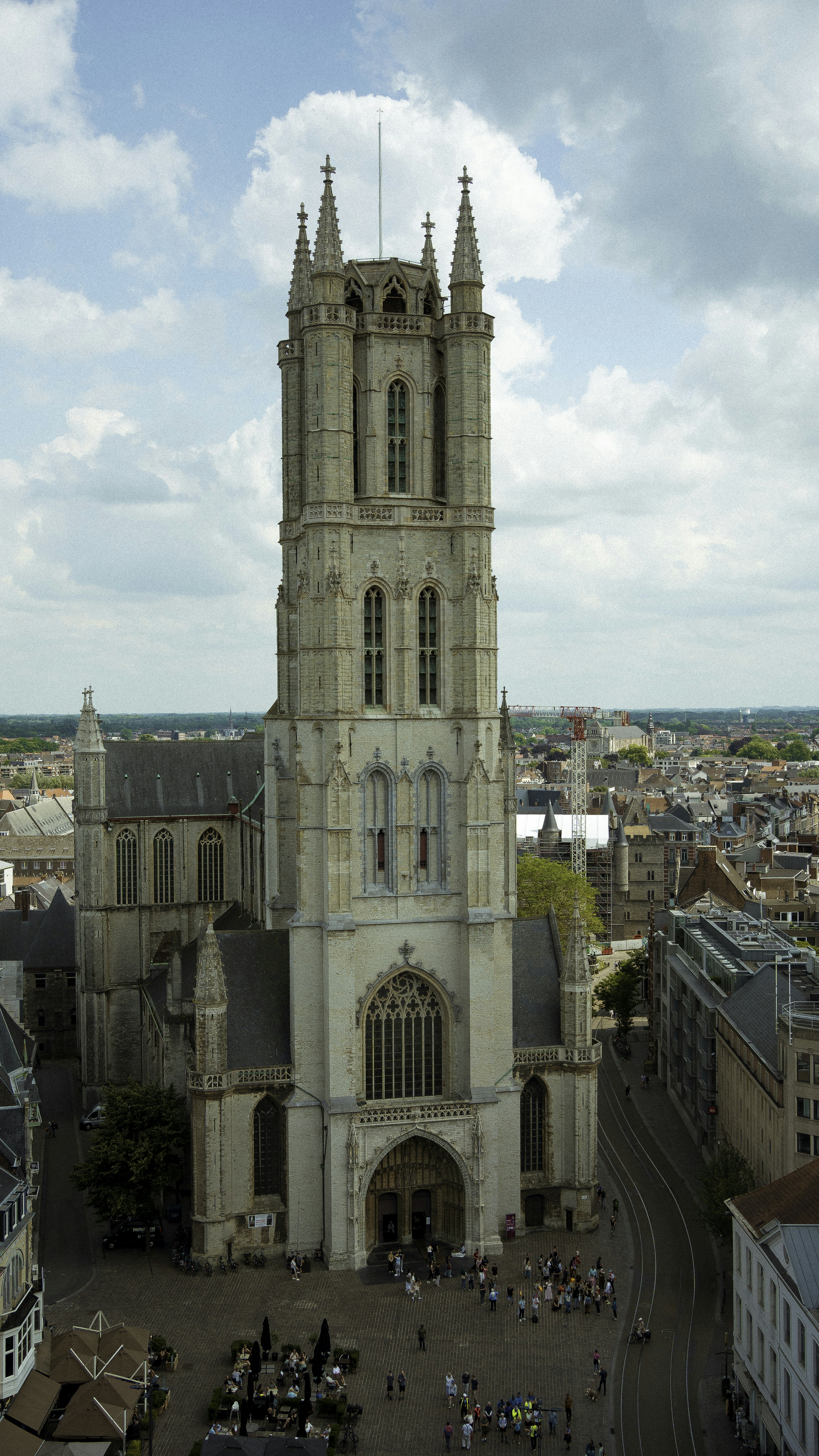 Gothic church tower rises above a bustling square, surrounded by historic buildings and lively crowds. The scene captures a blend of architectural grandeur and everyday life.