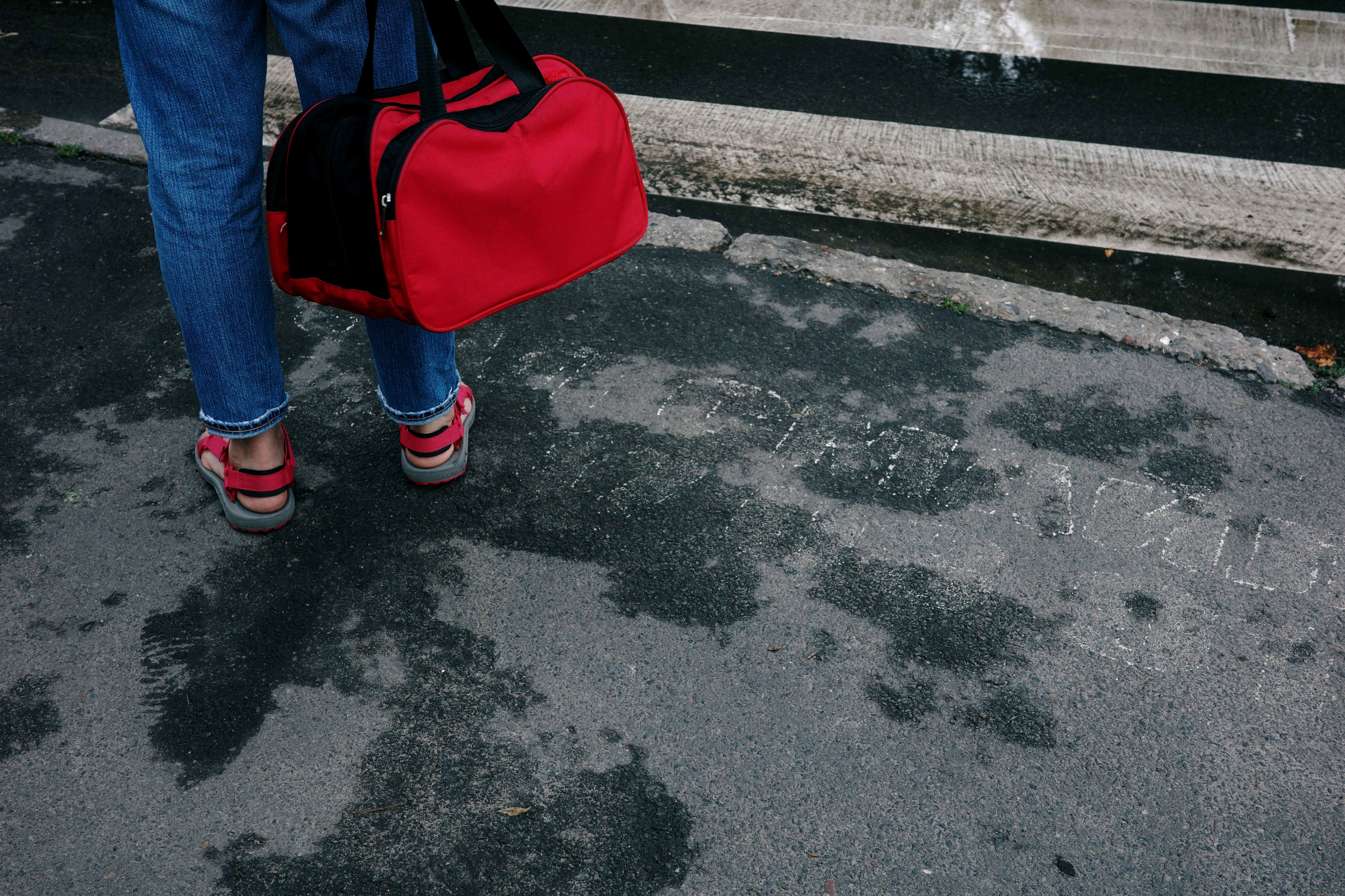 A person wearing blue jeans and red sandals walks on a wet, textured asphalt surface, carrying a vibrant red and black duffel bag. Behind them, a pedestrian crossing with distinct white stripes is visible. The ground shows patches of dampness, suggesting recent rain. The focus is on the lower half of the person's body and the ground, giving a sense of movement and an urban setting. | A person carries a red bag down the street.