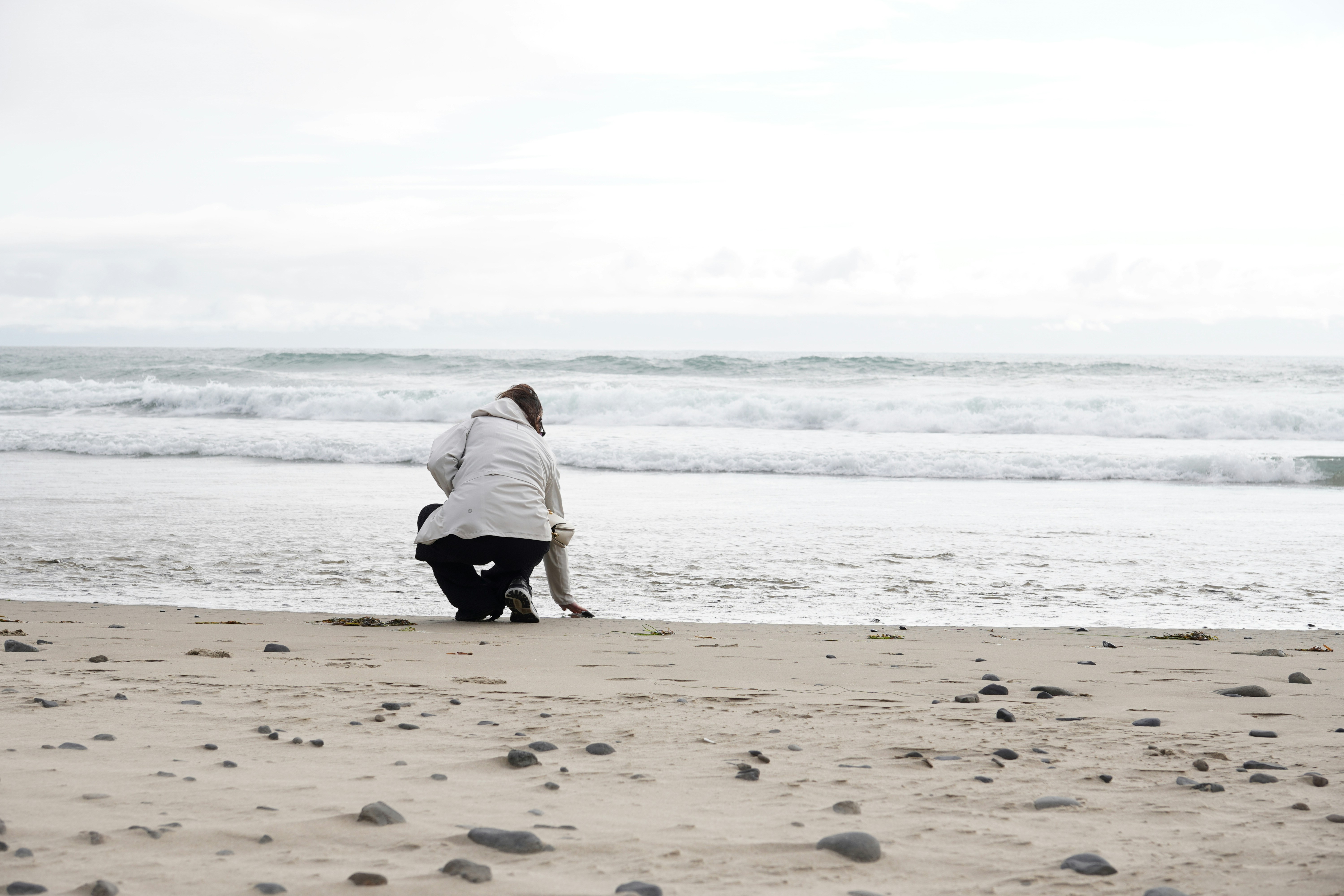 A woman in a white jacket crouches at the water’s edge, touching the incoming tide. With crashing waves and a cloudy horizon, the scene captures a quiet moment of connection to nature. The contrast of human presence and vast ocean adds emotional weight.