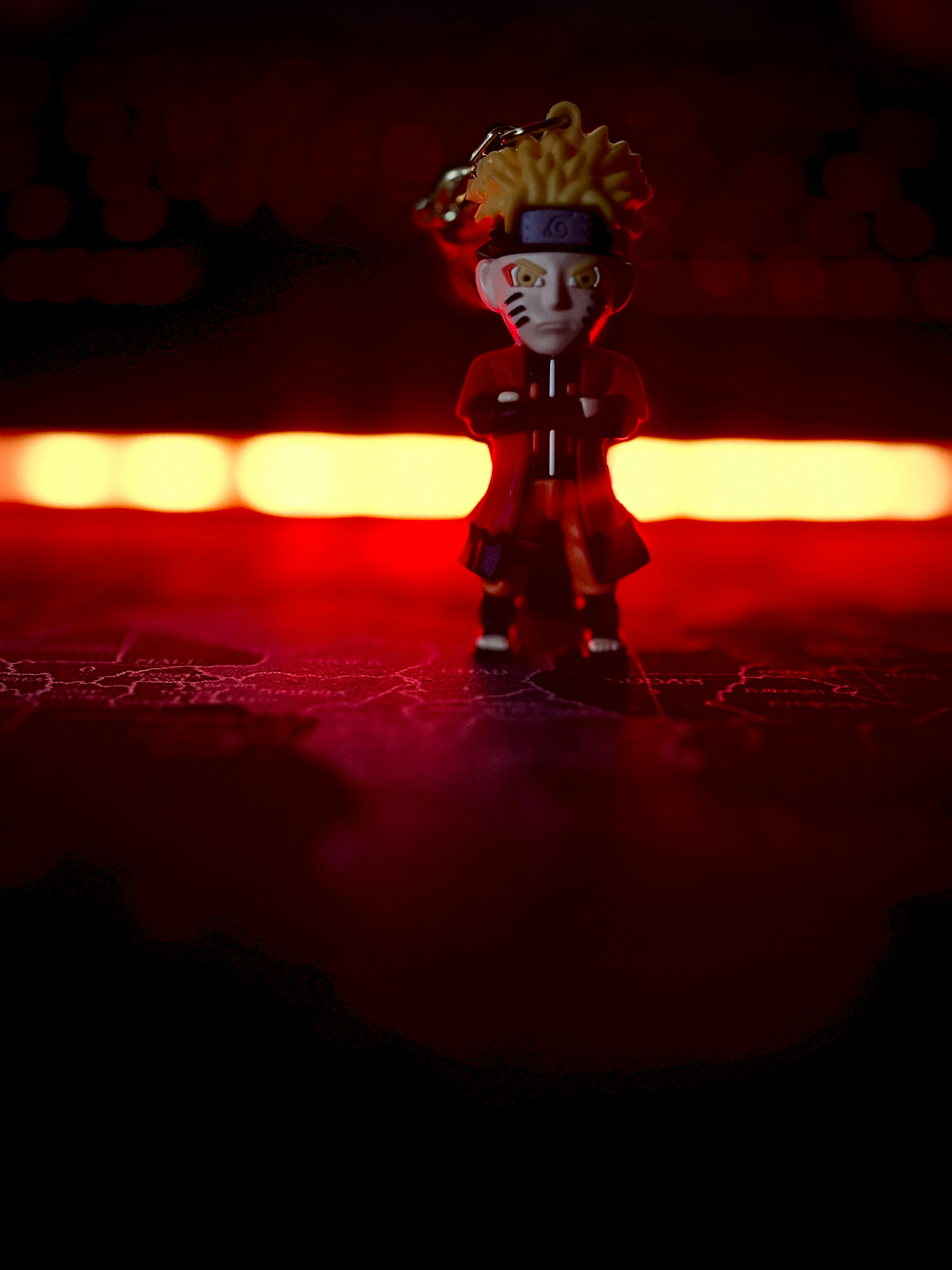 A striking close-up shot of a Naruto Uzumaki keychain figure captured in dramatic low lighting. The figure stands with arms crossed, bathed in an intense red glow that creates a powerful, fiery atmosphere. The blurred background and illuminated surface give the photo a cinematic, almost battle-ready feel — perfectly echoing Naruto’s fierce determination and iconic presence. | Naruto stands in front of a red lit background.
