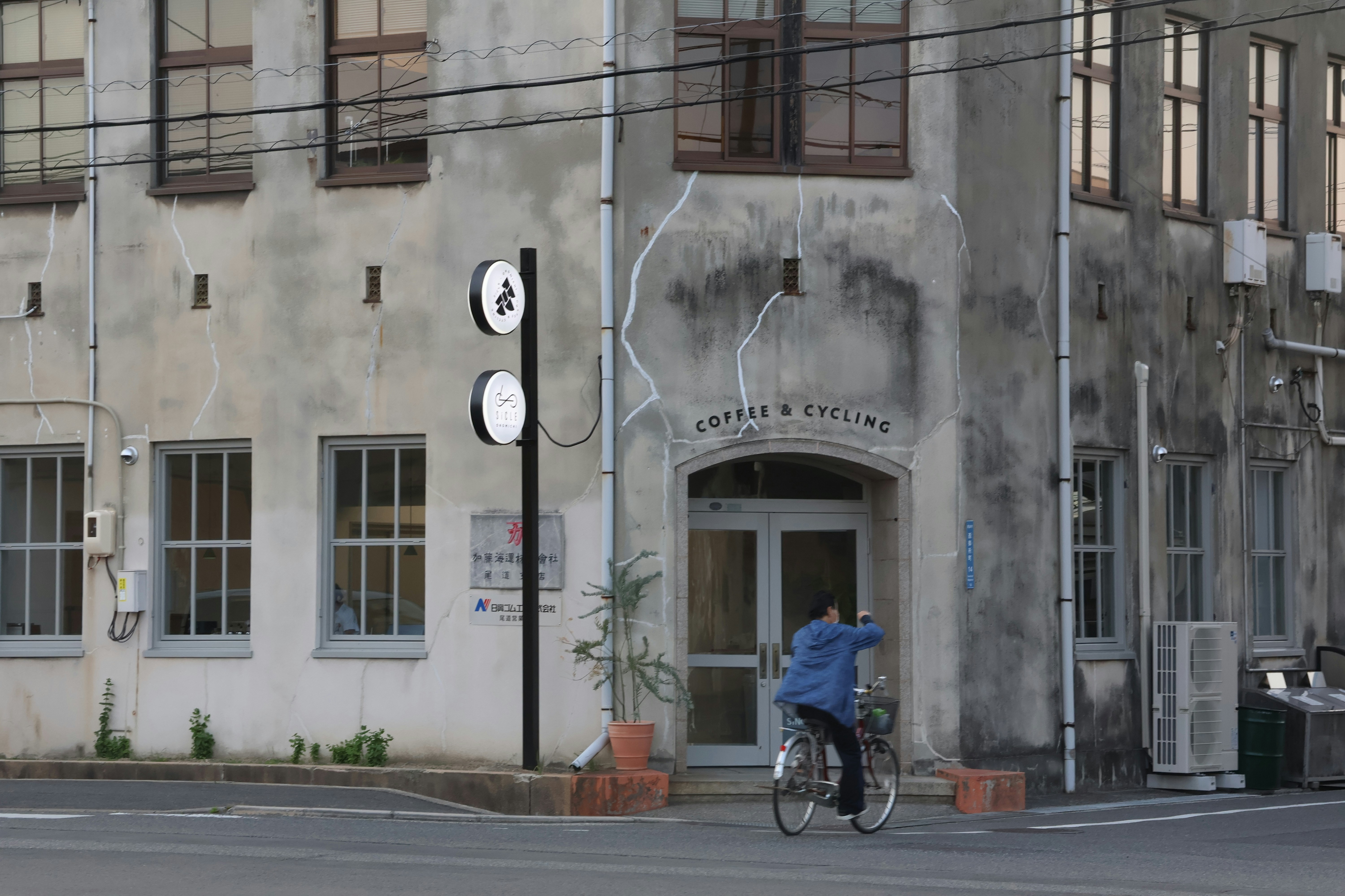 A cyclist passes by a quaint coffee shop and cycling hub, showcasing a blend of urban life and leisure in a weathered building. The scene reflects a tranquil moment in a bustling city.